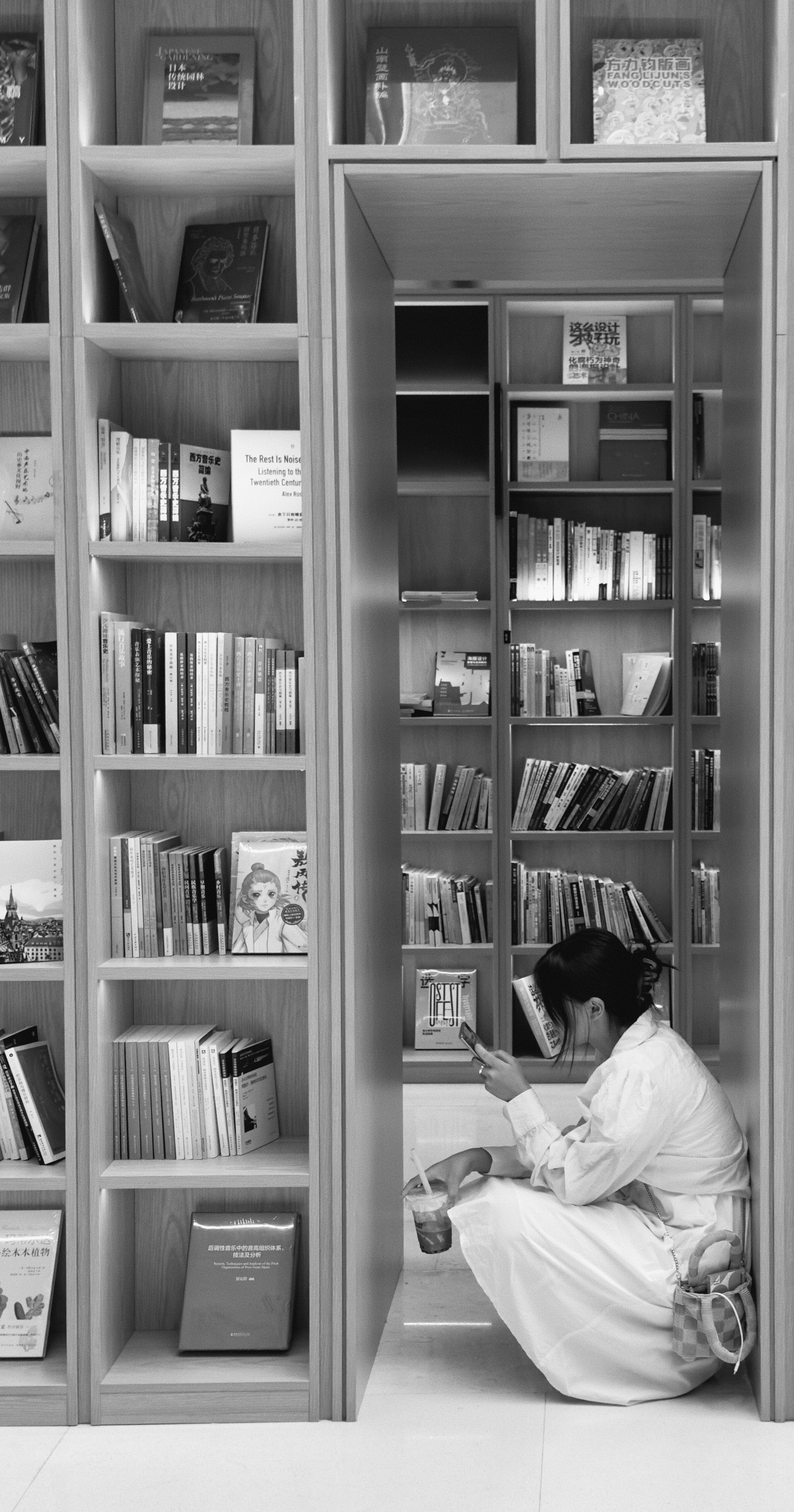 For some people, their days are nothing more than crossing off names one by one on a to-do list, never fully immersing themselves in life — it's like watching rain through a glass window: you know it's raining, but you can't feel the coolness of the rain or smell the earthy scent when raindrops hit the soil. We're always chasing efficiency, rushing through days faster than a tram, yet forgetting to stop and take a good look at what's right in front of us. The pressure to get things done is like a whip, lashing us forward, leaving no time to even think about other possibilities. However, only b | Woman sitting in a bookshelf nook with a drink.