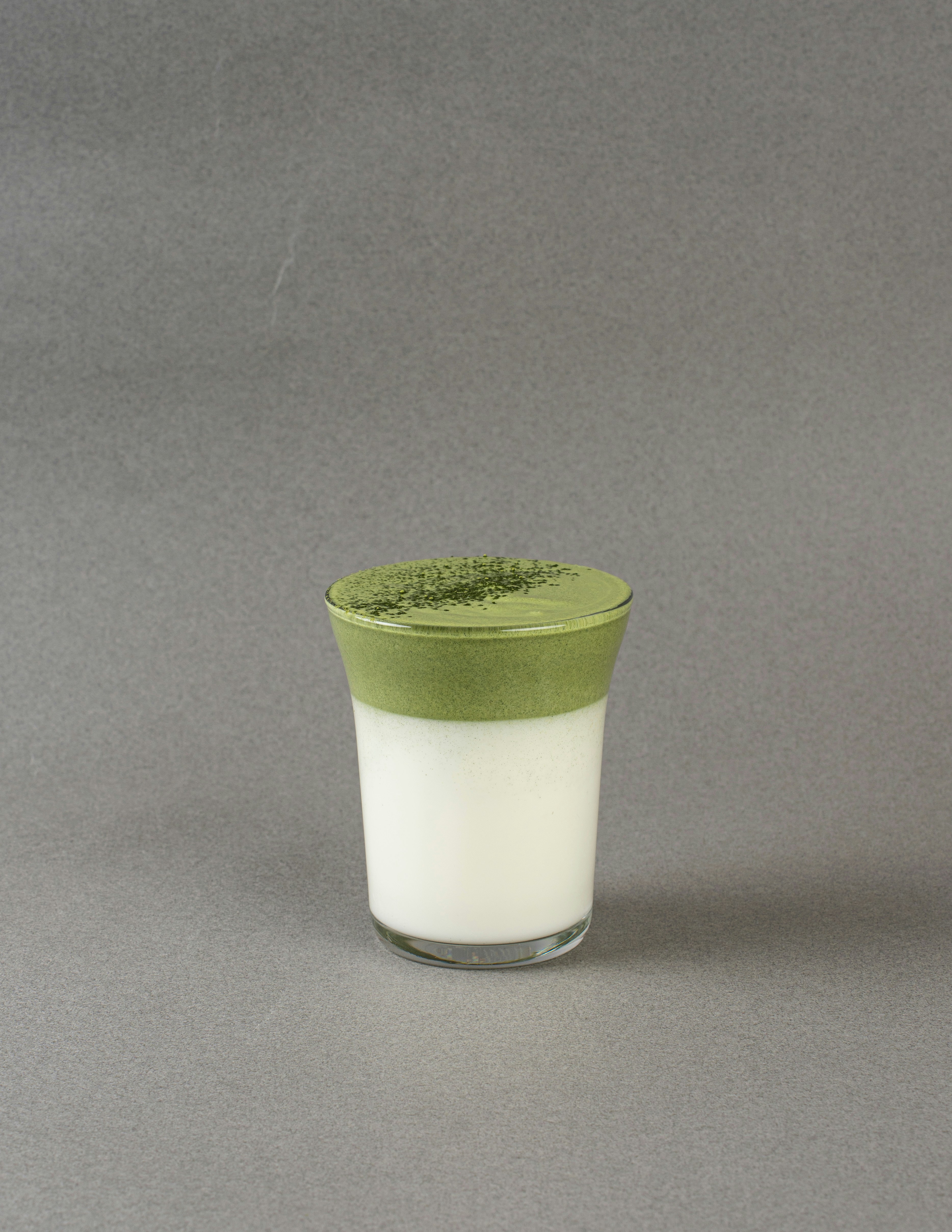 A delicate glass filled with creamy matcha topped with a green froth, set against a muted gray backdrop.