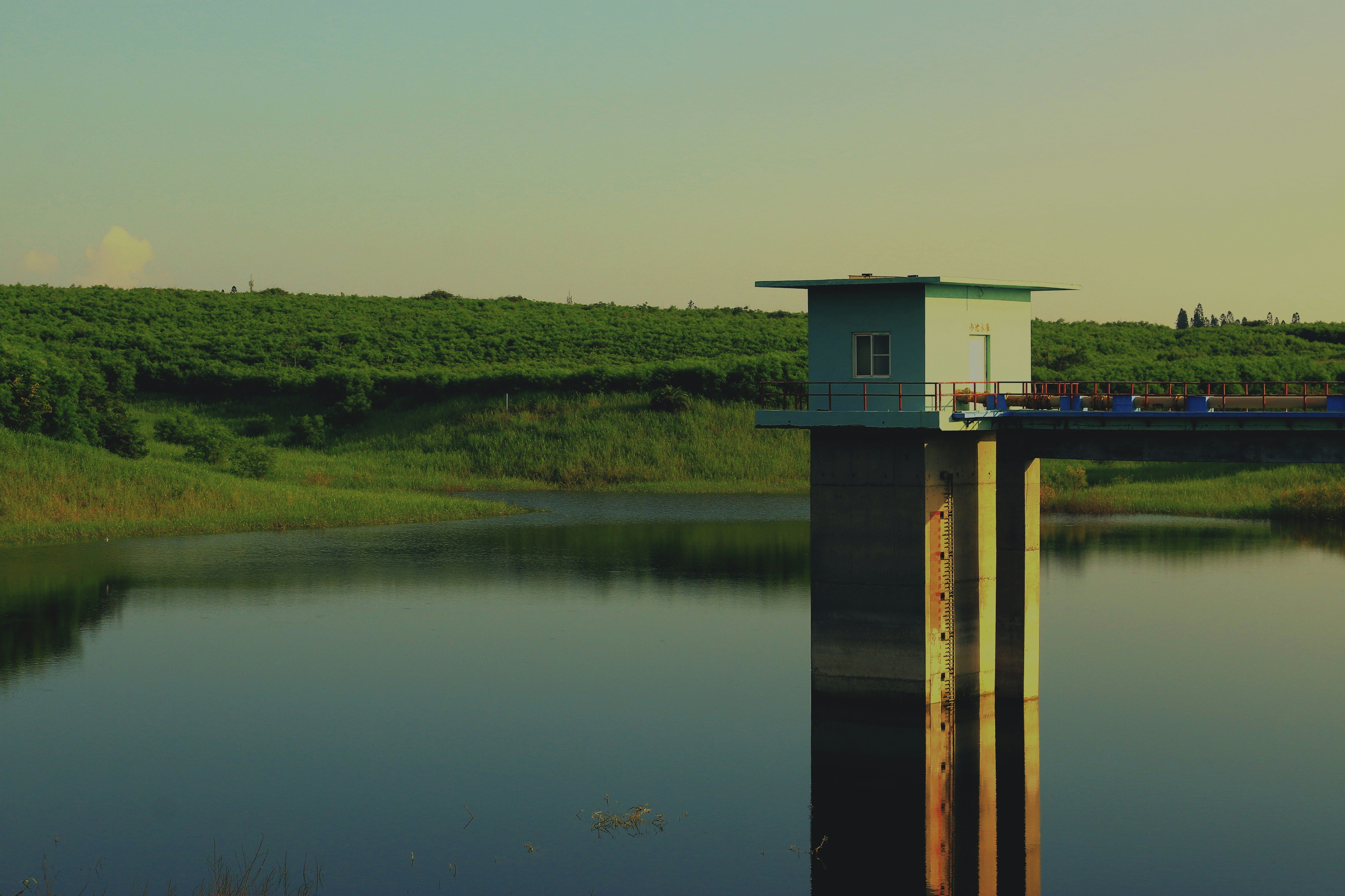 A solitary control tower stands over a tranquil body of water, surrounded by lush greenery under a soft sky.