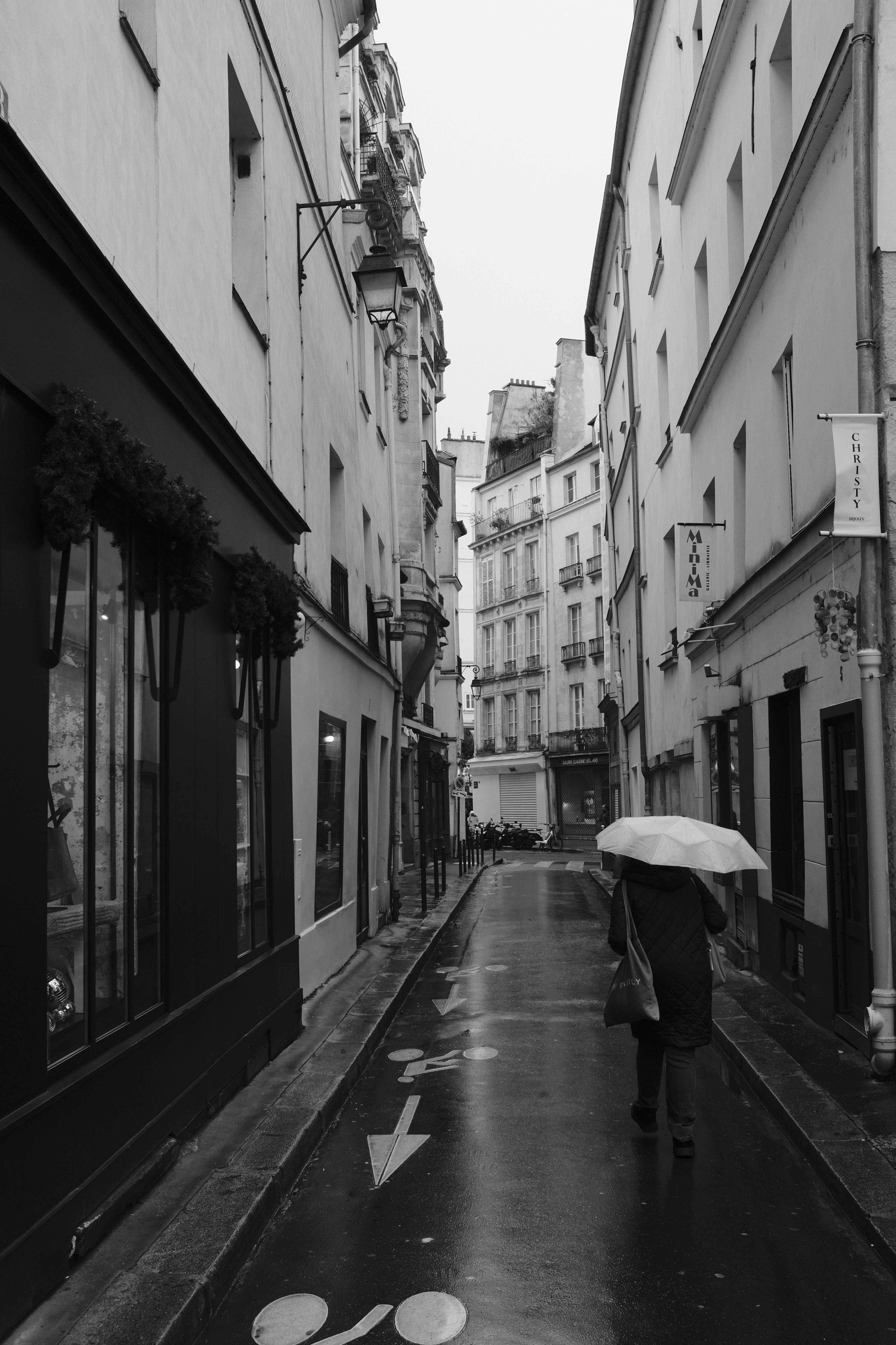 A lone figure walks down a narrow, rain-soaked street in Paris, flanked by charming buildings and soft street lamps. The atmosphere is serene and reflective.