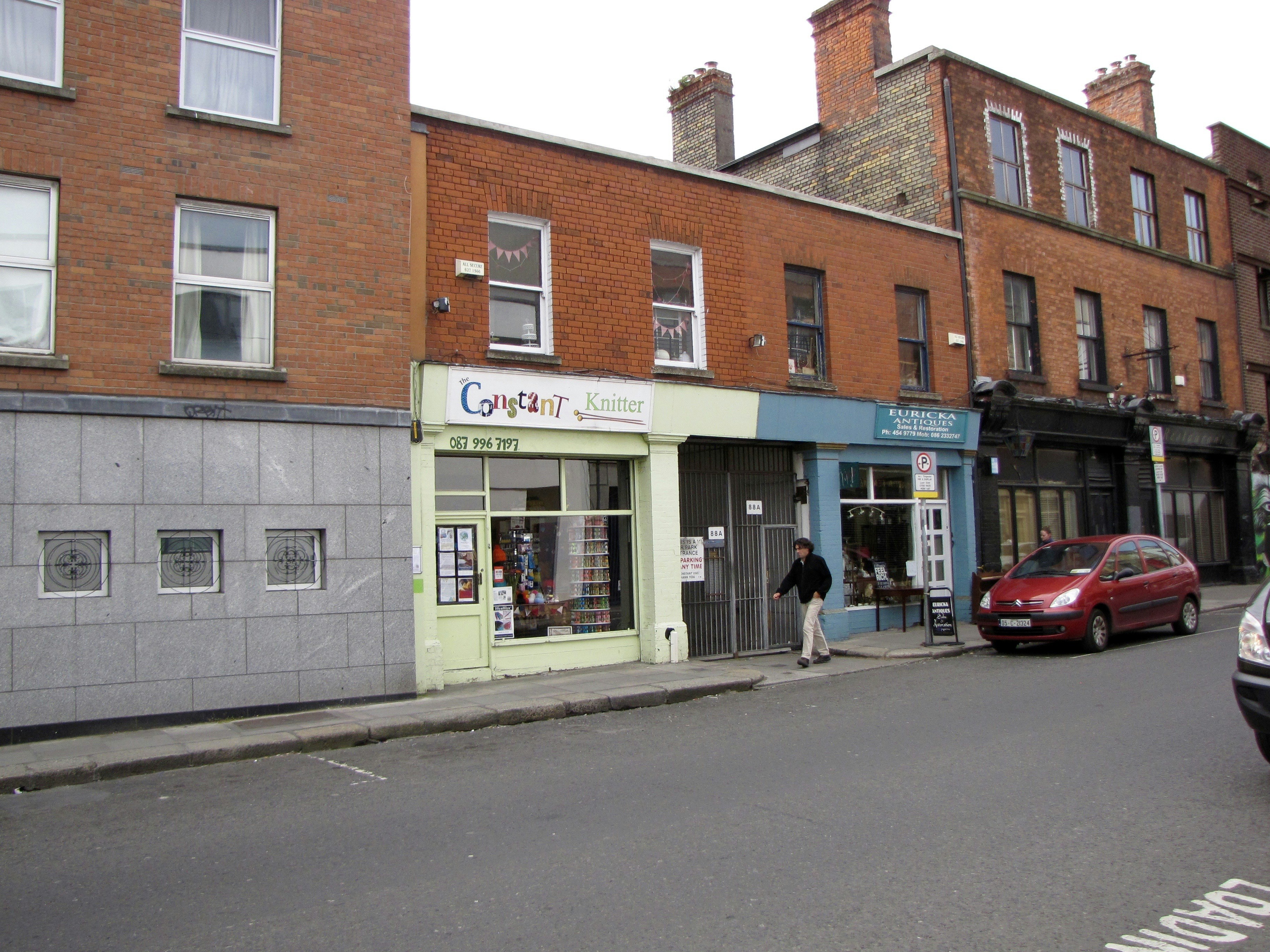 Colorful storefronts line a quiet street, with a pedestrian passing by and a parked car nearby.