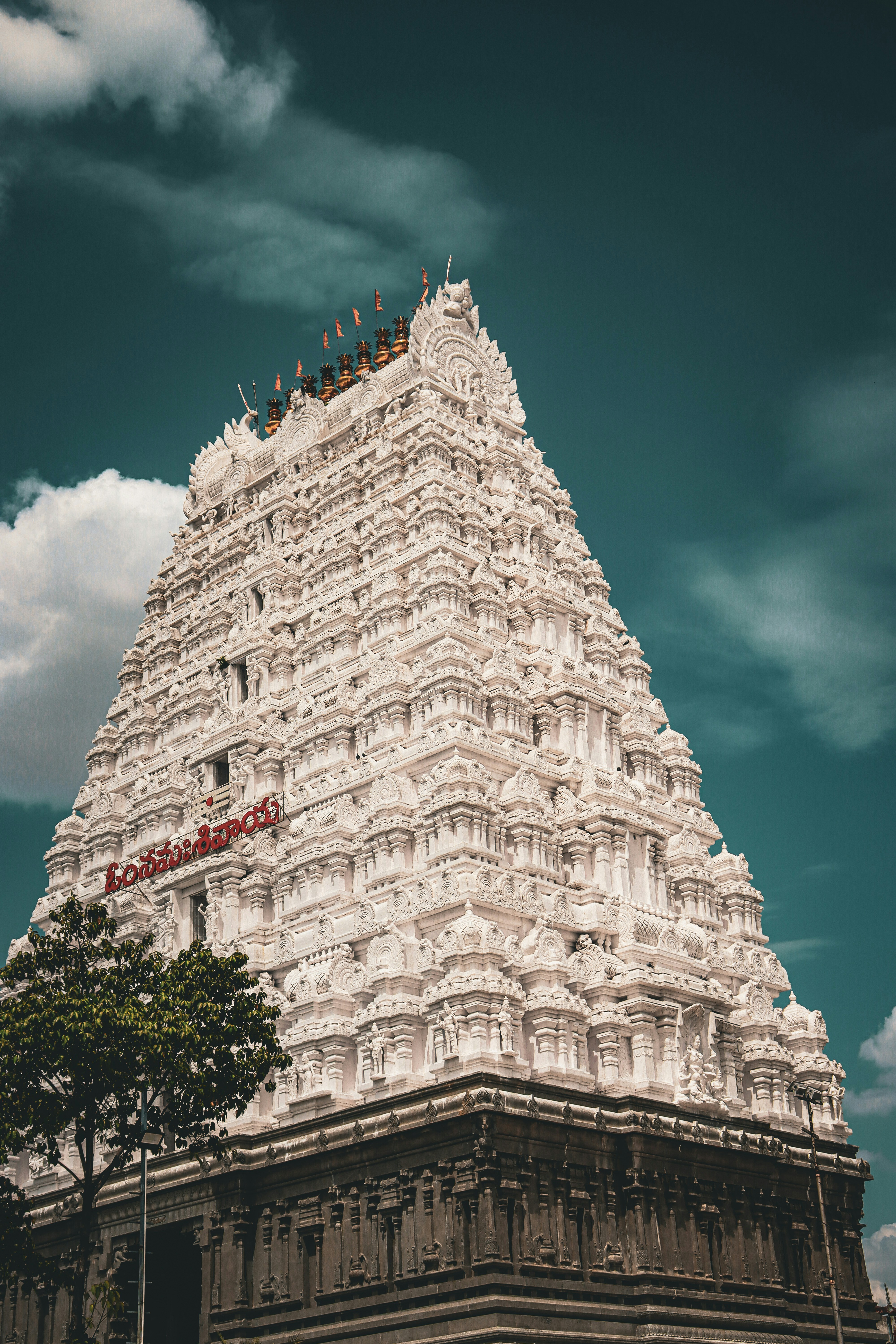 Intricate white temple tower adorned with detailed carvings, set against a dramatic sky. The vibrant red inscription adds a striking contrast.
