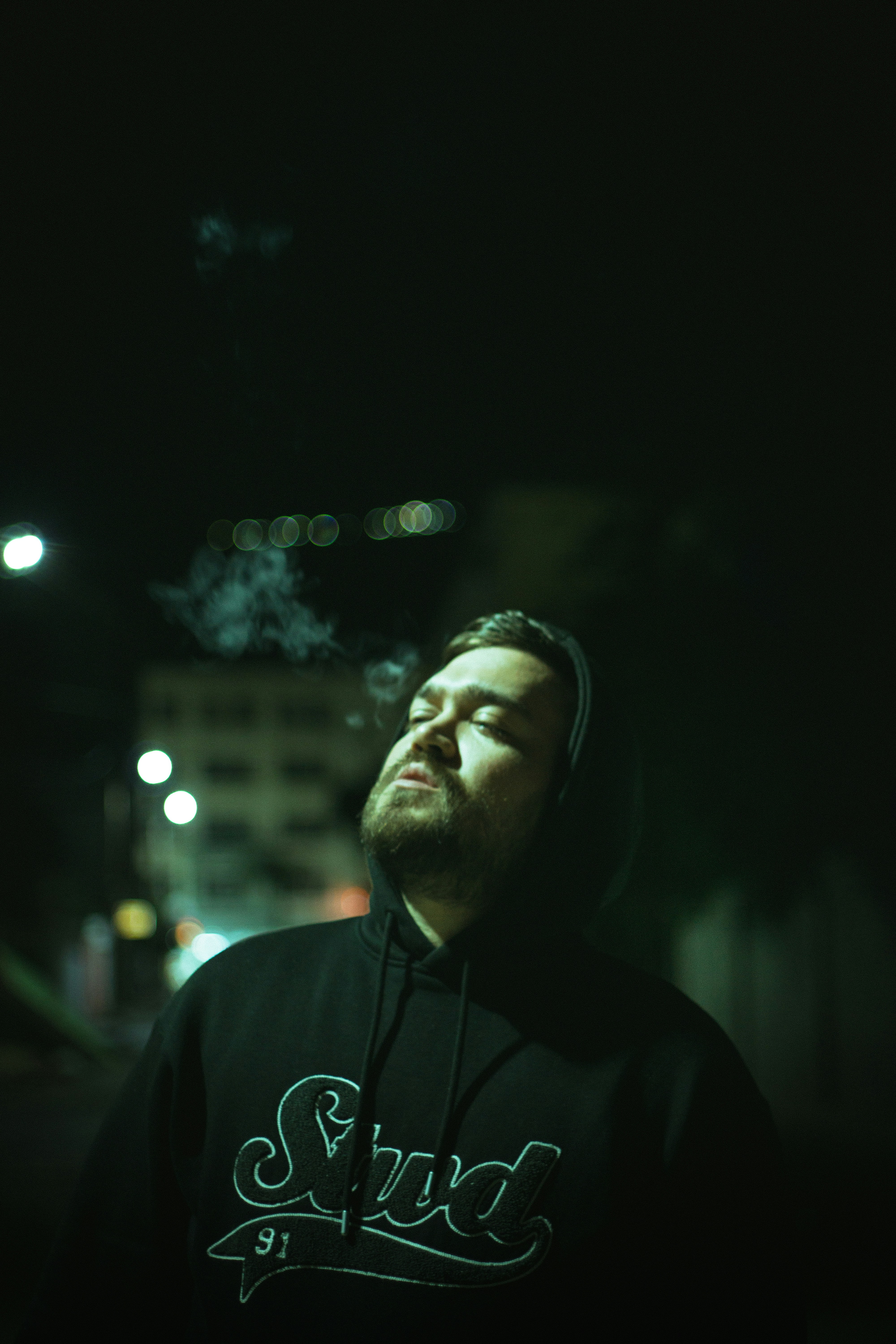 Man in a hoodie standing in a dimly lit urban street, eyes closed, seemingly lost in thought. Soft lights from the background create a moody atmosphere.