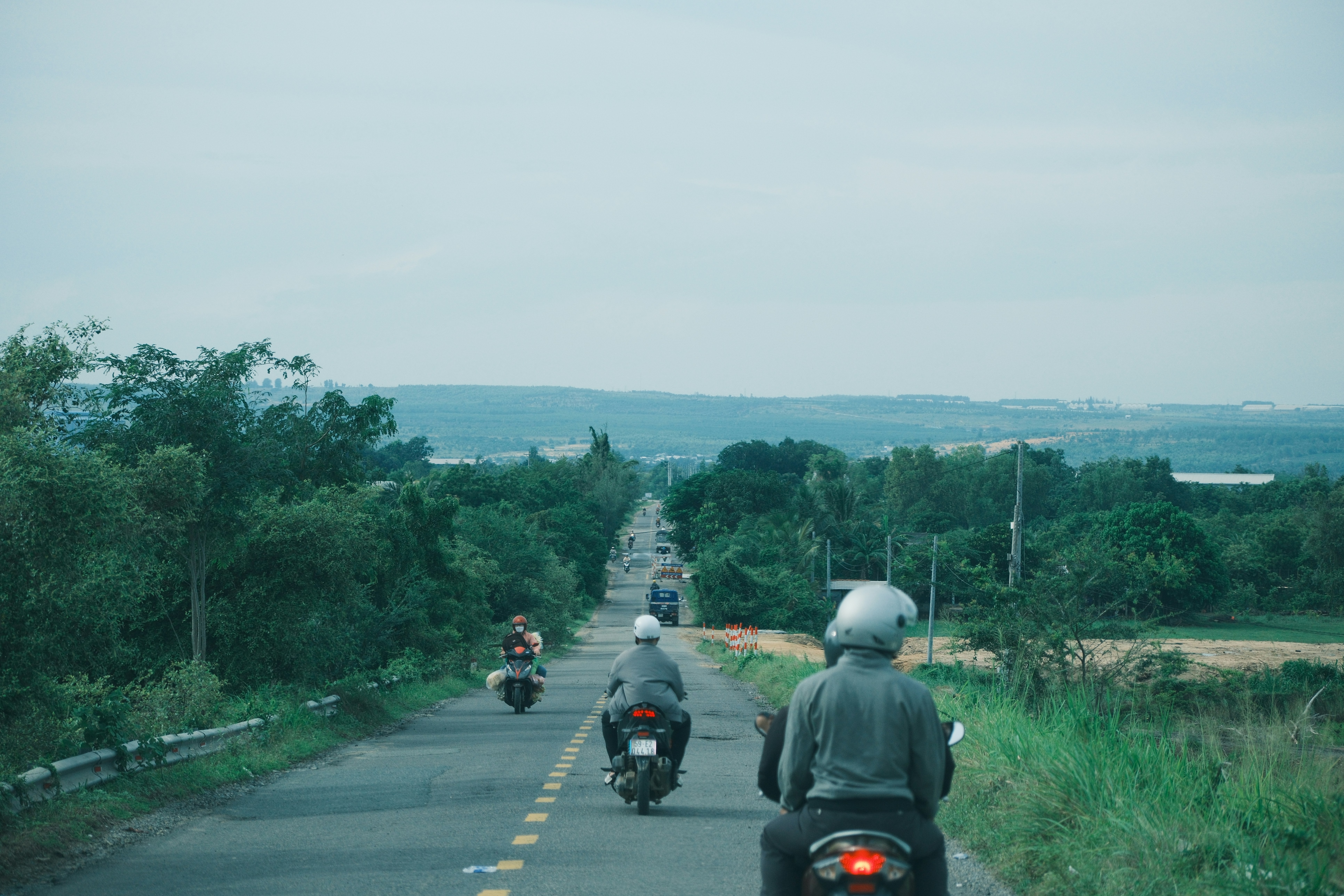 Motorcyclists navigating a winding road flanked by lush greenery, leading towards distant hills under a cloudy sky.