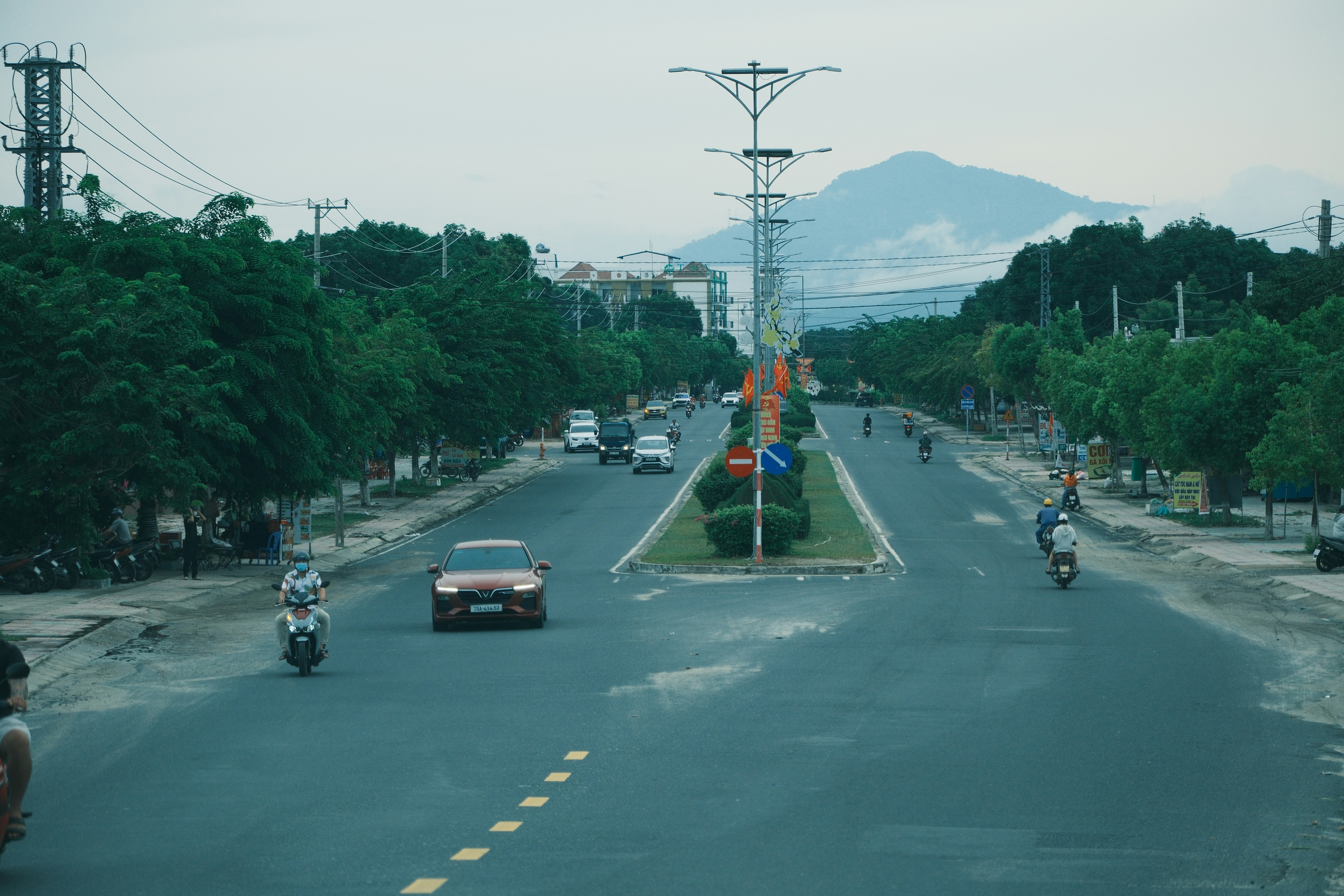 A bustling street scene showcasing vehicles and lush trees lining the road, with distant mountains providing a serene backdrop.
