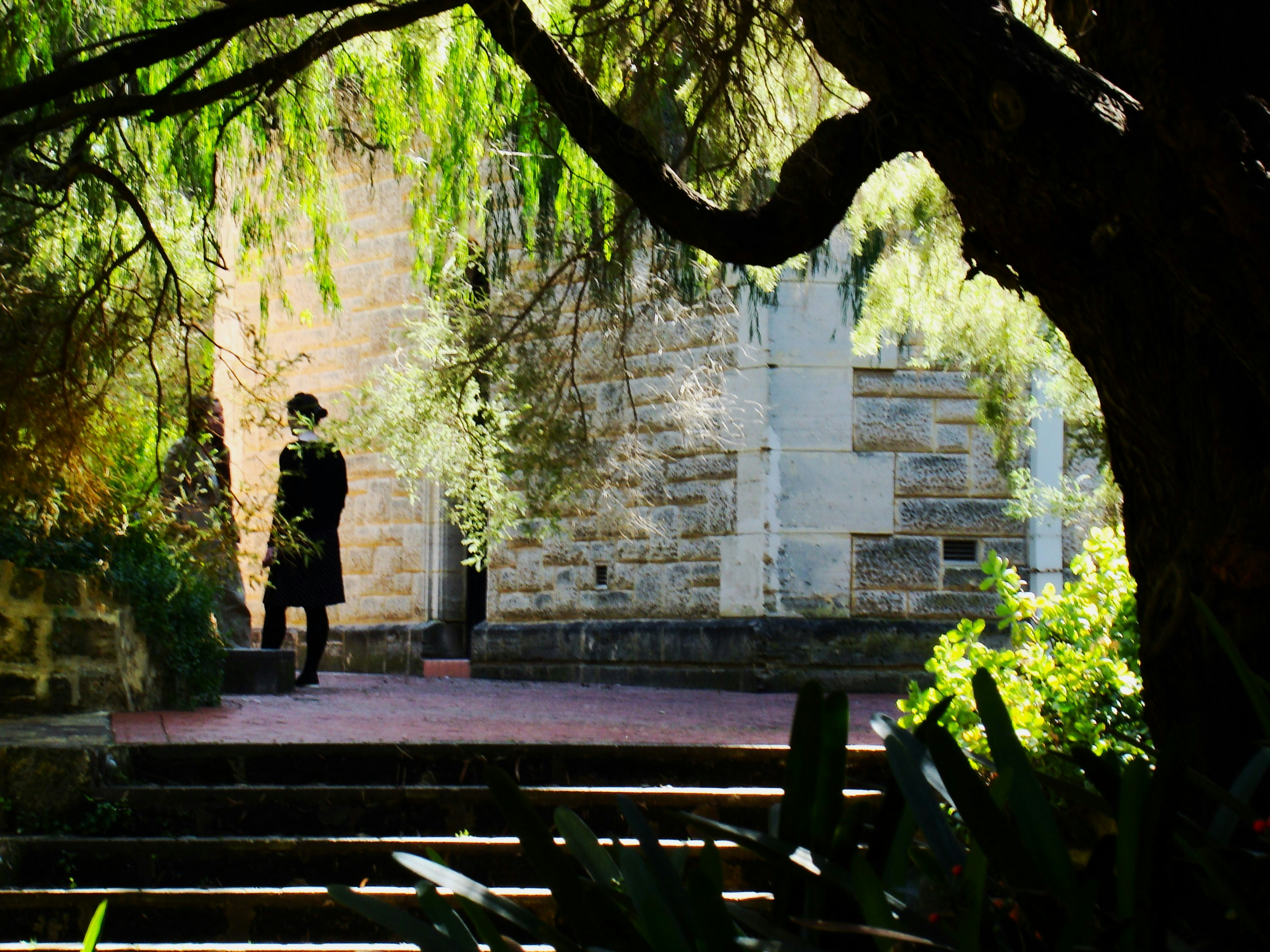 Person in hat stands near stone building in garden.
