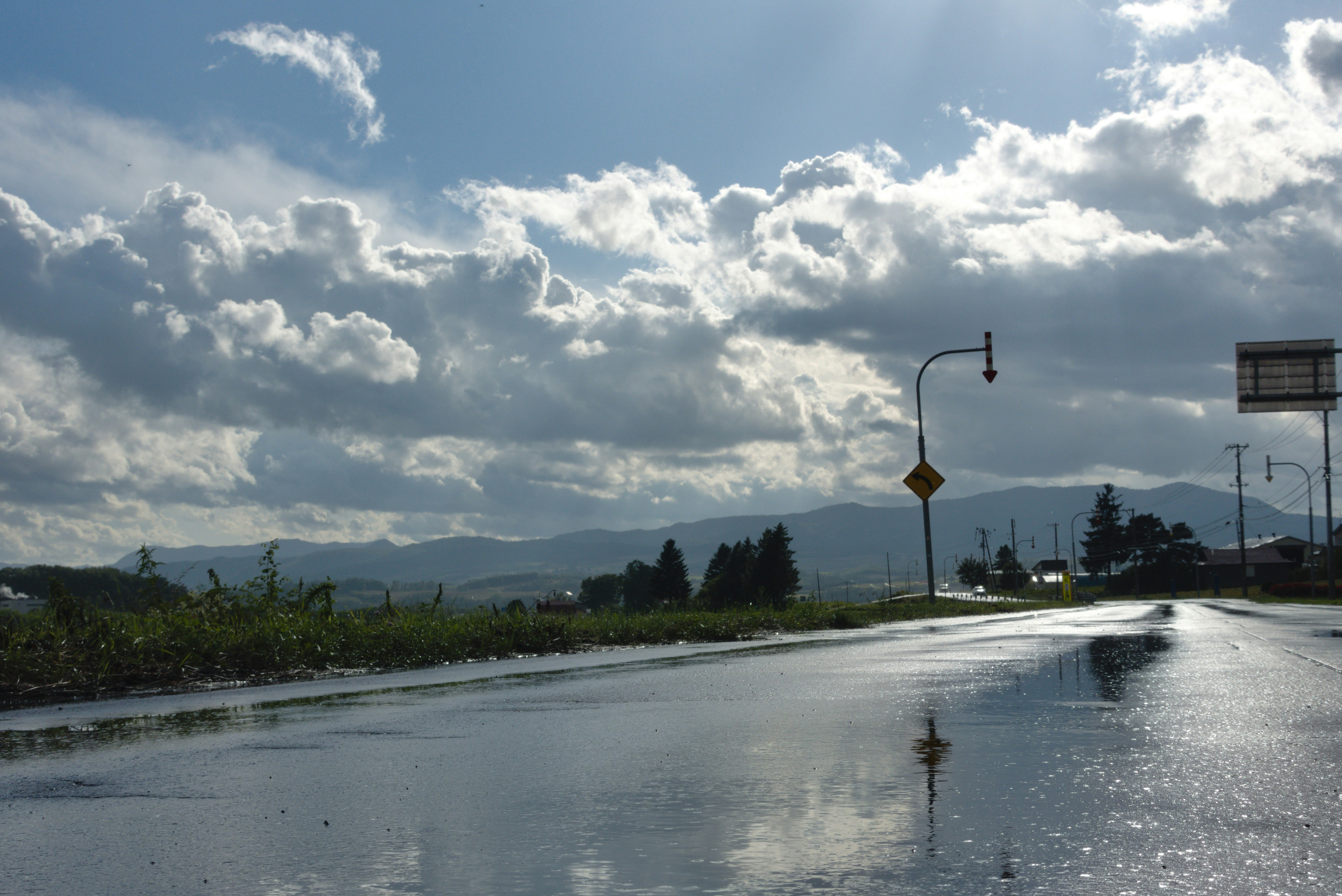Wet road reflecting clouds and distant mountains under a dramatic sky. A traffic sign and pole add perspective to the scene.