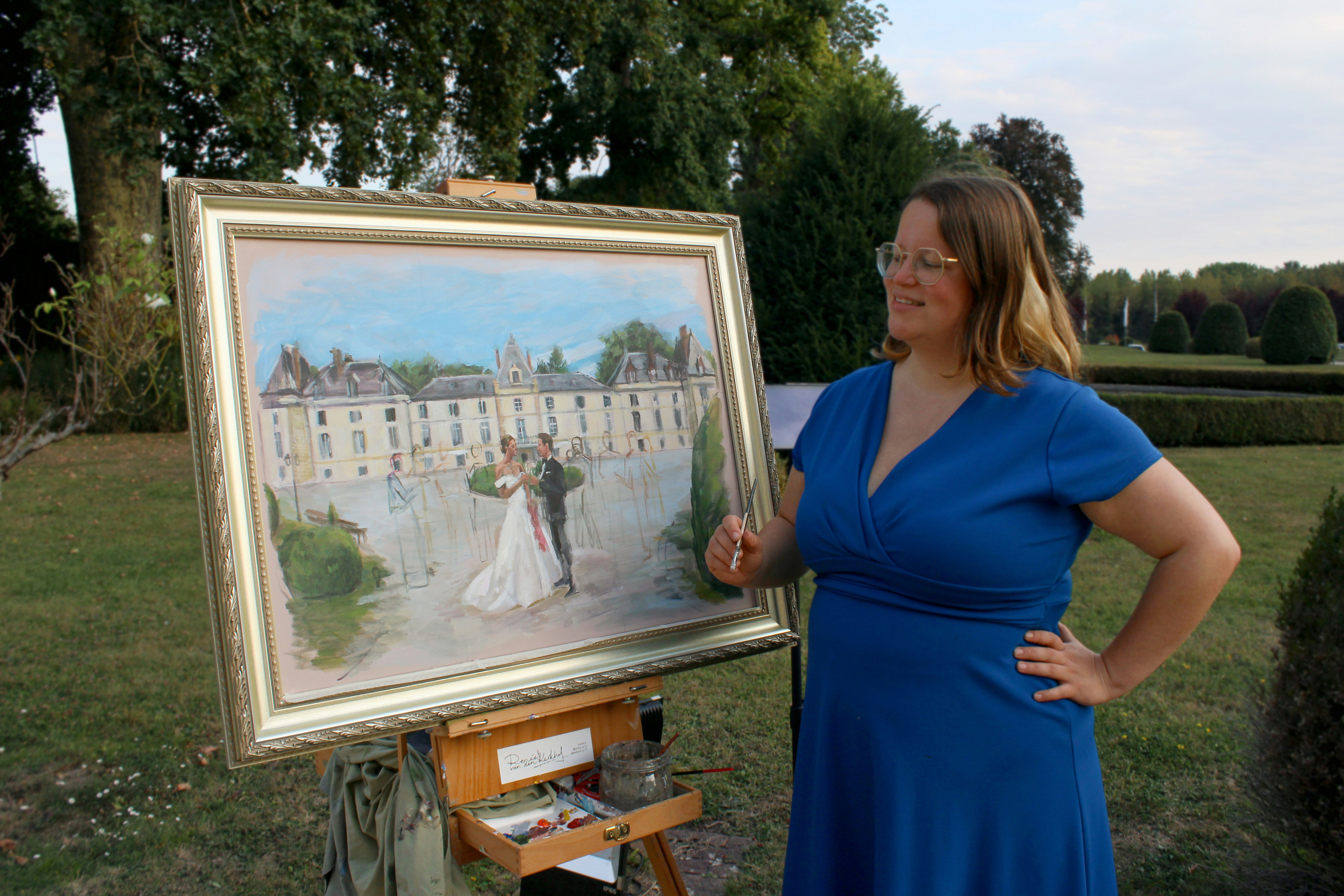 Destination wedding in France: Dutch live painter Renée wearing a blue maxi dress, painting an outdoor wedding scene on location with the couple in the center. The setting of the artwork is in front of Château D'aveny, a lovely, luxury 18th century castle. The artist is working with a paintbrush and a foldable French box easel - in the background we see the castle gardens. | Woman in blue dress paints a wedding couple portrait.