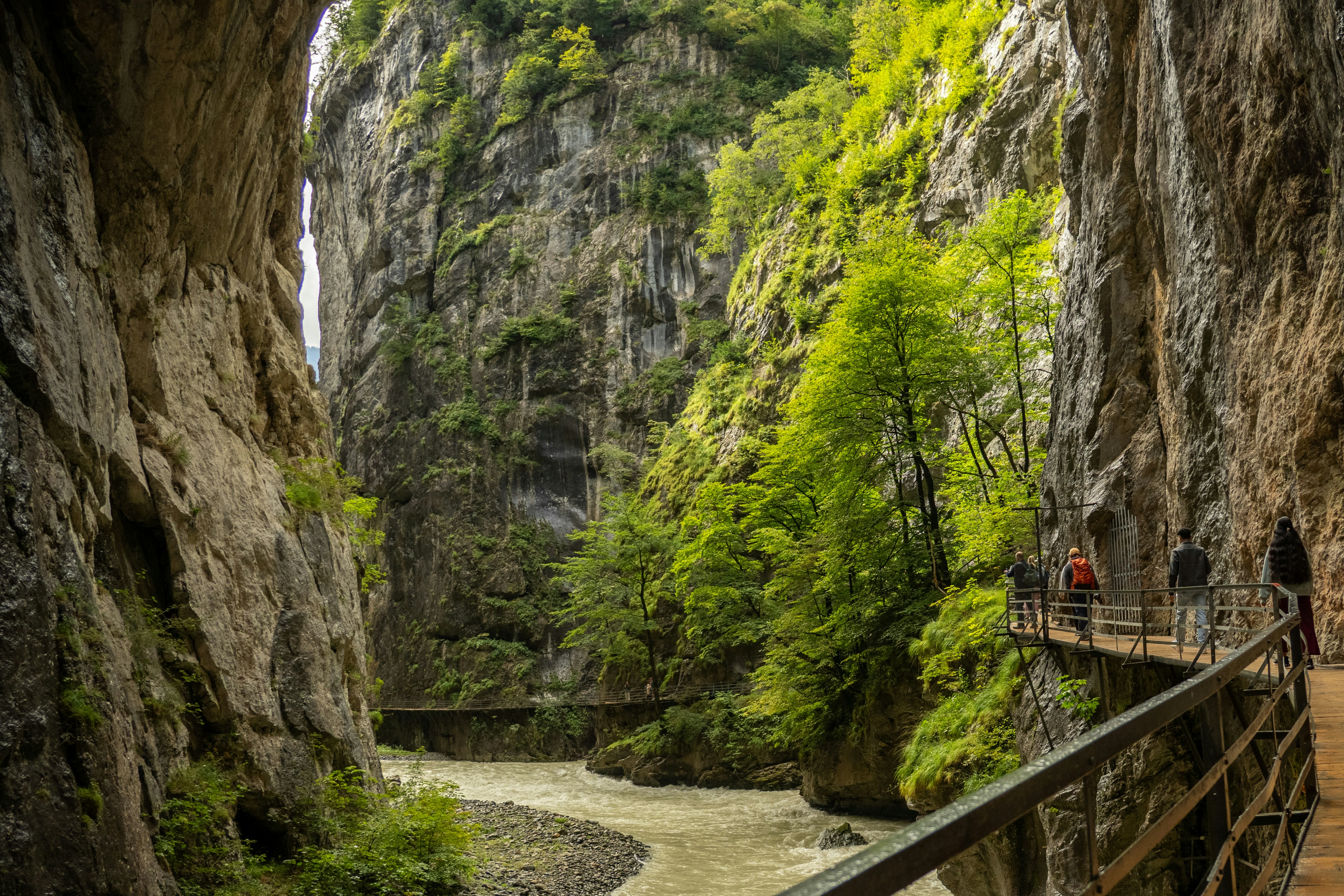 A view of a river and a wooden walkway in a narrow, rocky gorge. | Hikers on a path beside a river in a canyon