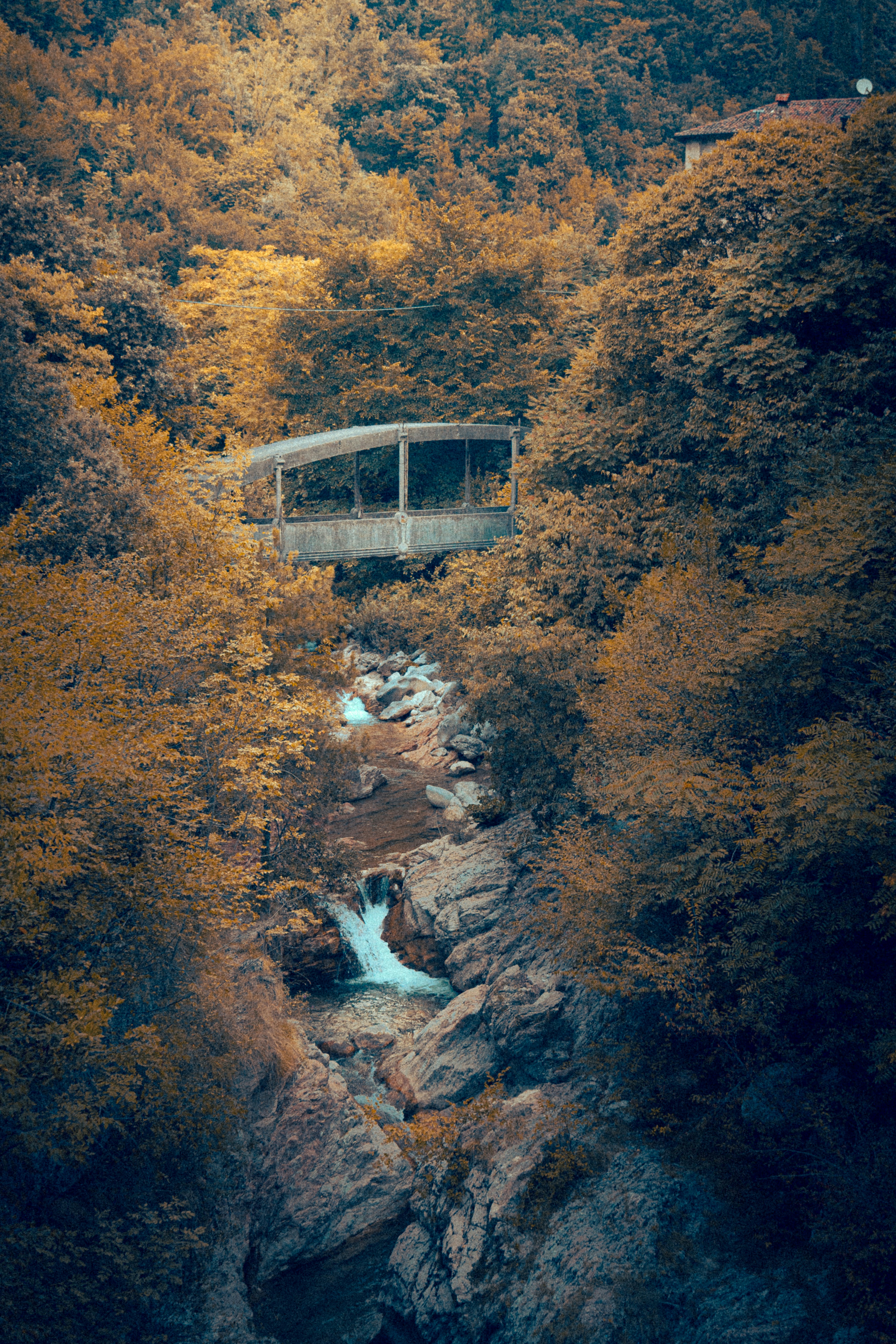 A stone bridge spans a rocky creek enveloped by autumnal trees in warm orange tones, viewed through a narrow valley.