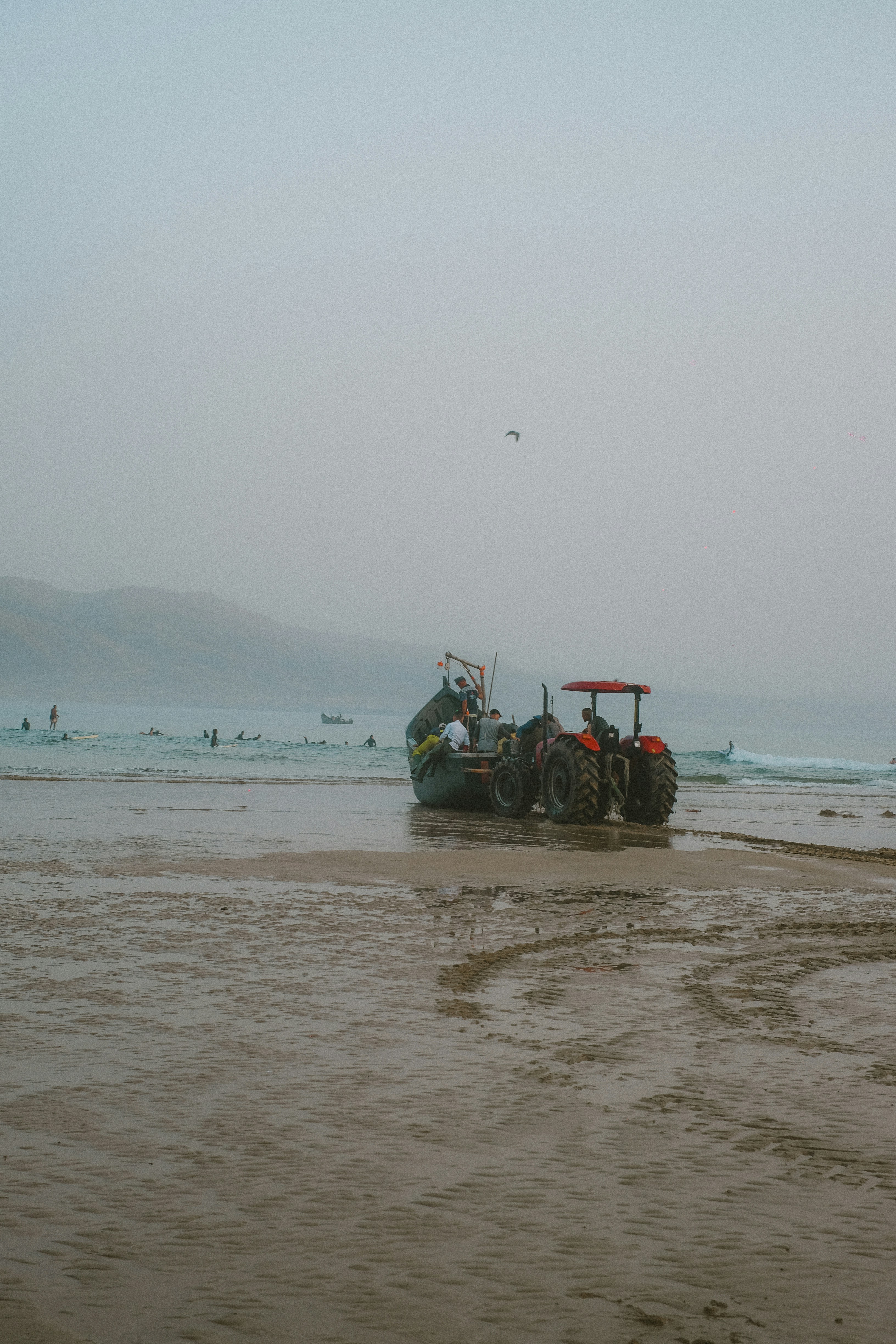 A tractor and fishing boat sit on the wet sand as surfers ride waves in the background under a foggy sky.