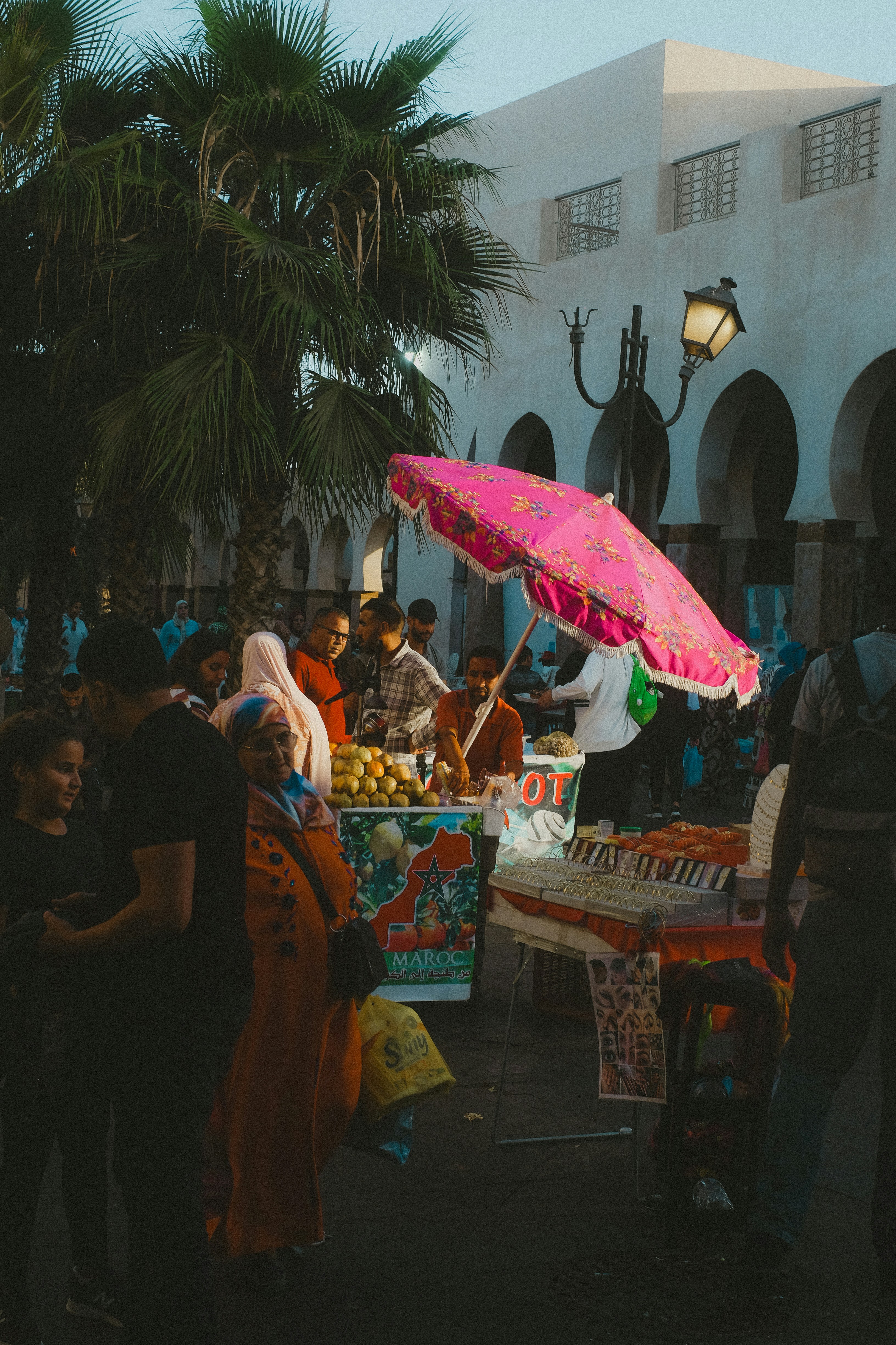 Bustling market with vendors and shoppers, featuring a colorful umbrella and fresh produce. The lively atmosphere captures the essence of community life.