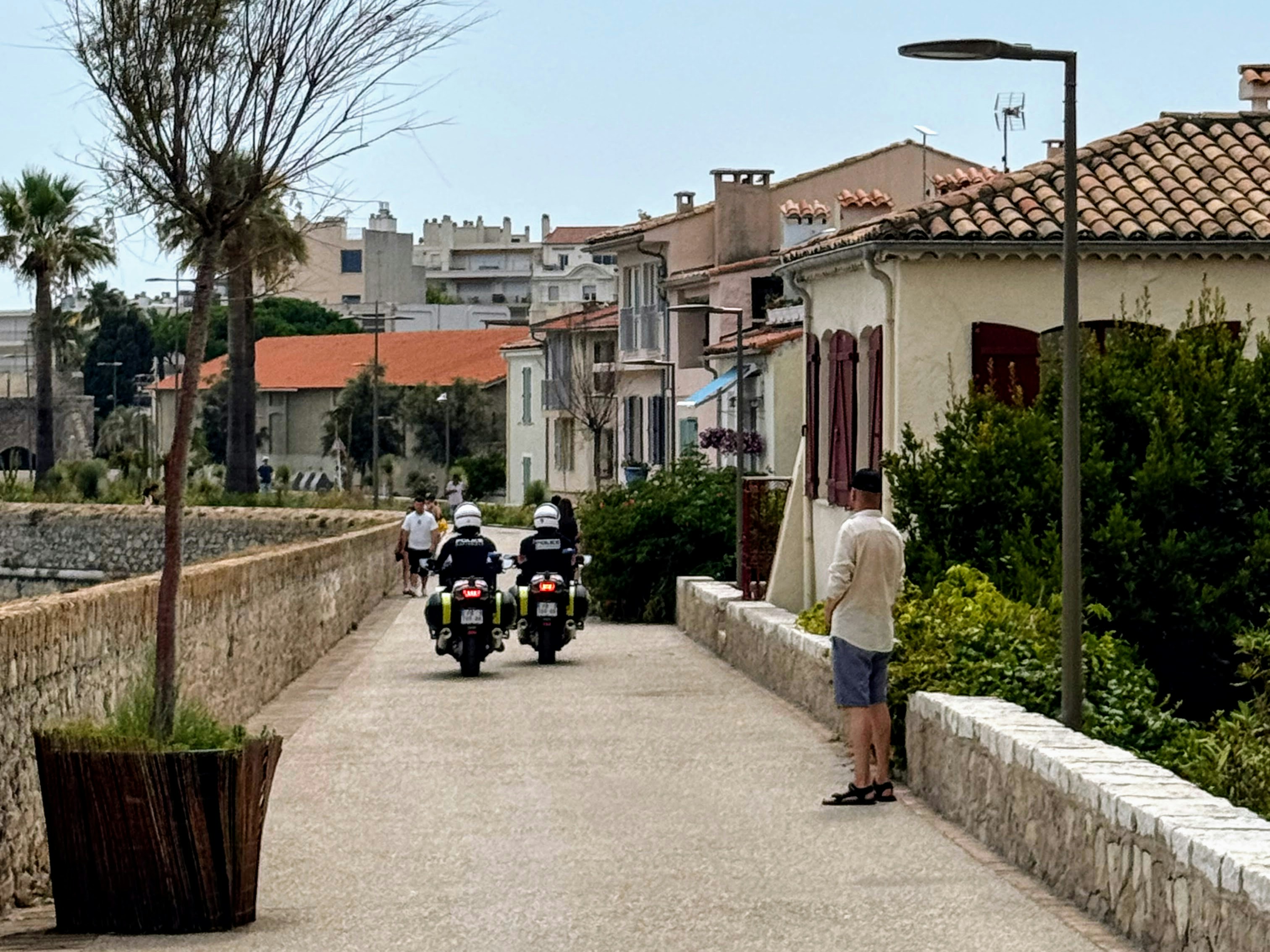 Two police motorcycles parked along a coastal walkway, with a pedestrian enjoying the view amidst charming houses and greenery.
