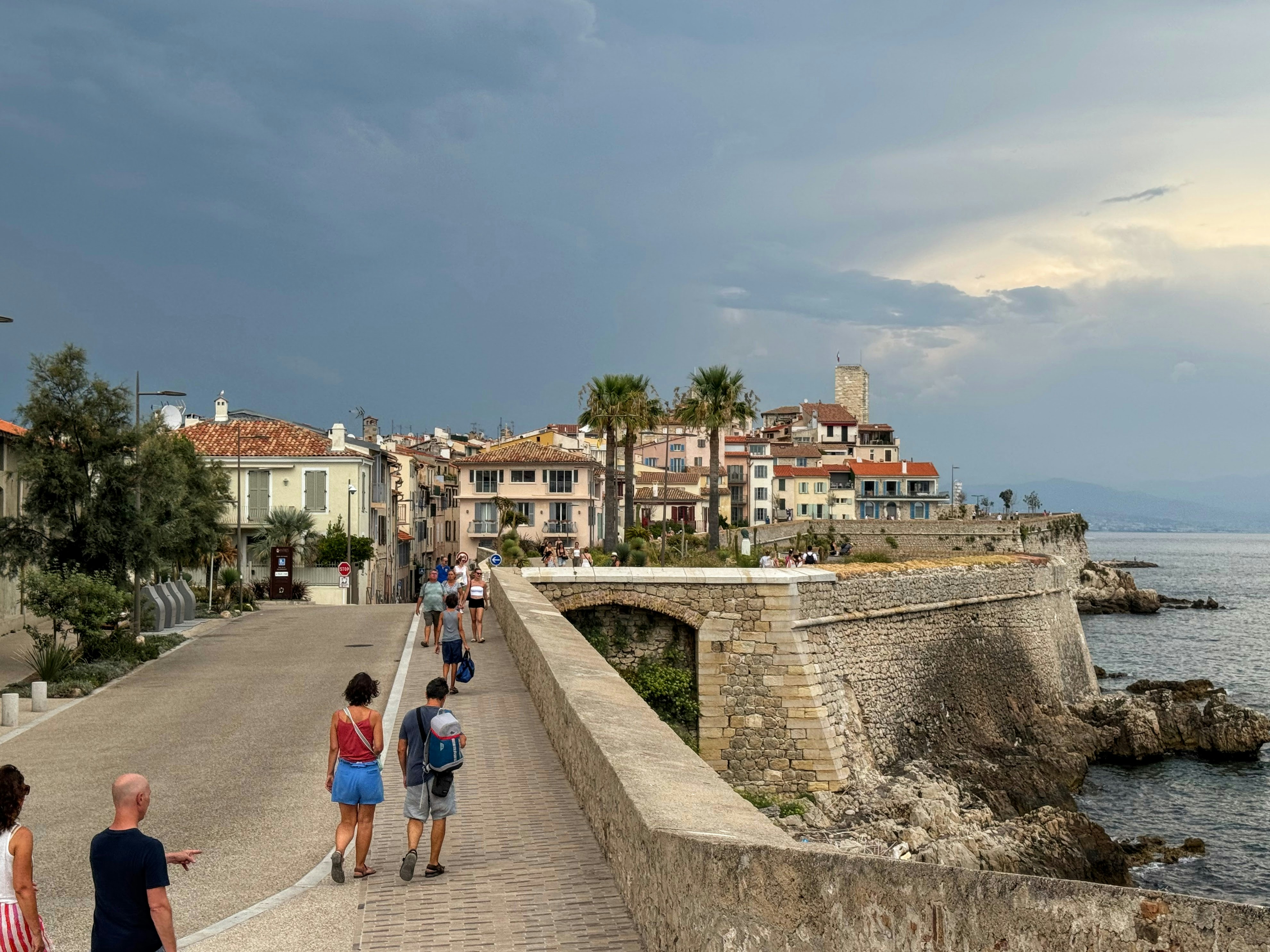 Pedestrians leisurely walking along a coastal path lined with charming buildings and palm trees, under a moody sky.