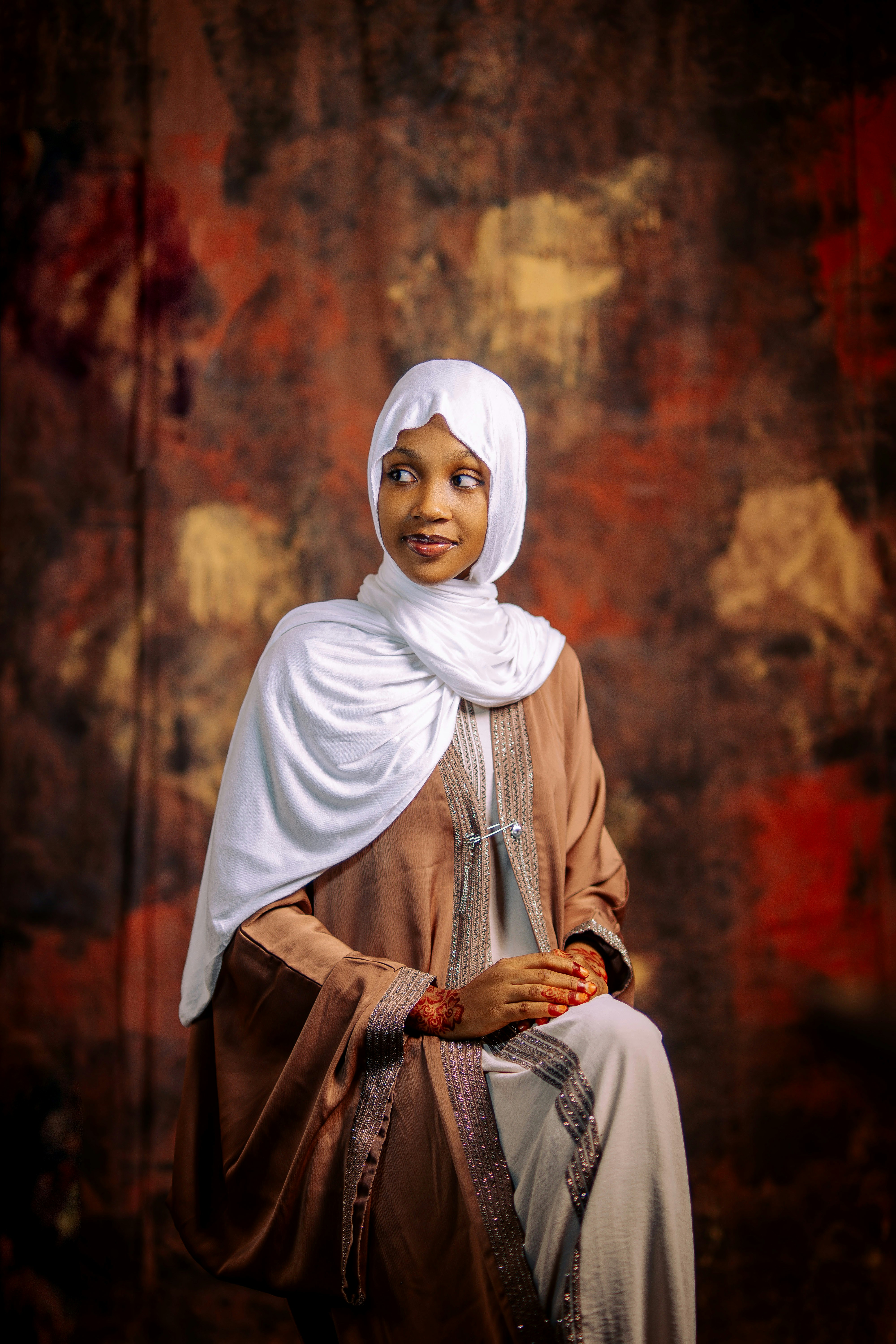 A young woman in traditional attire, adorned with a white hijab, poses gracefully against a textured, colorful backdrop.