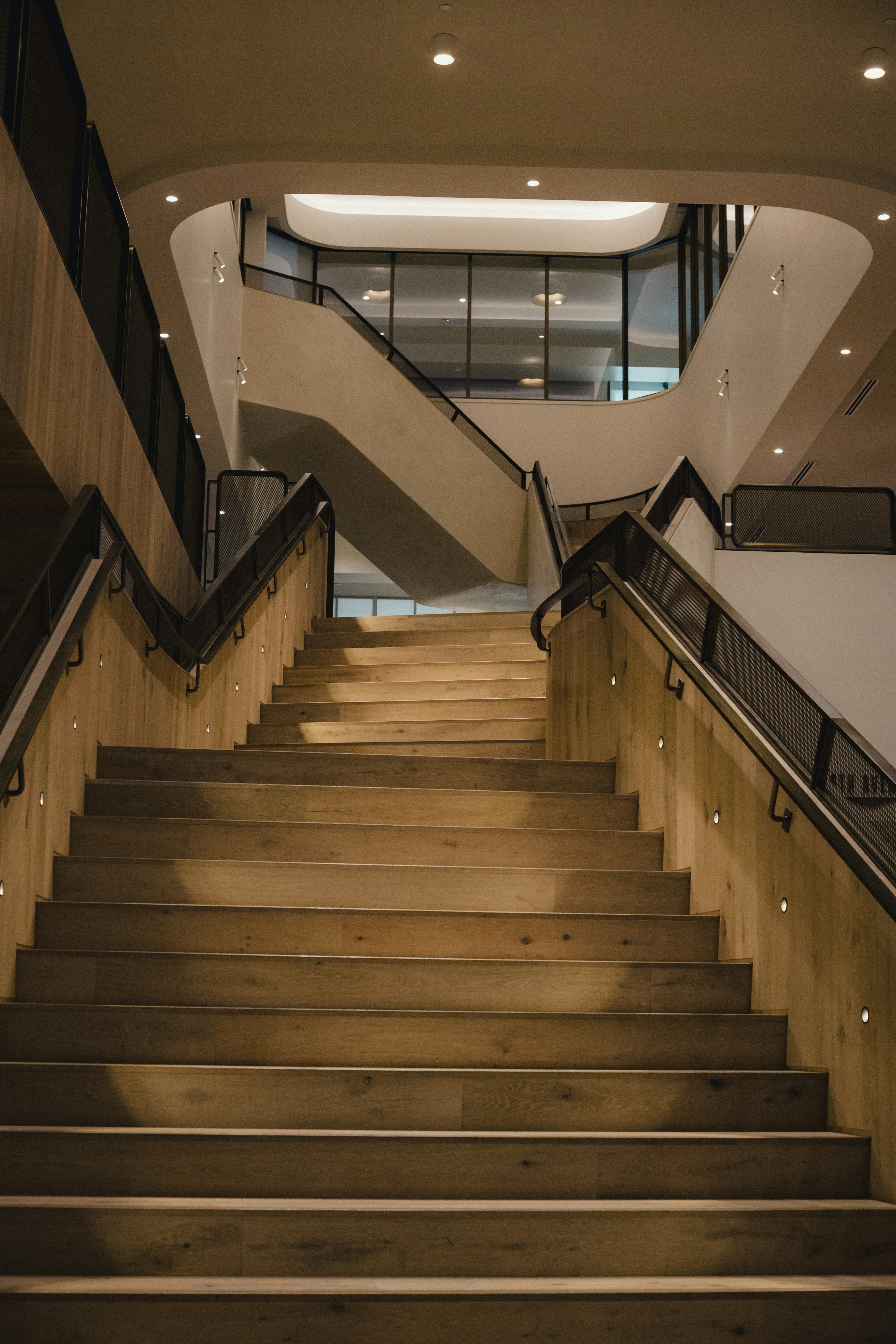 Elegant wooden staircase leading to a spacious upper level, illuminated by soft lighting and surrounded by contemporary architecture.