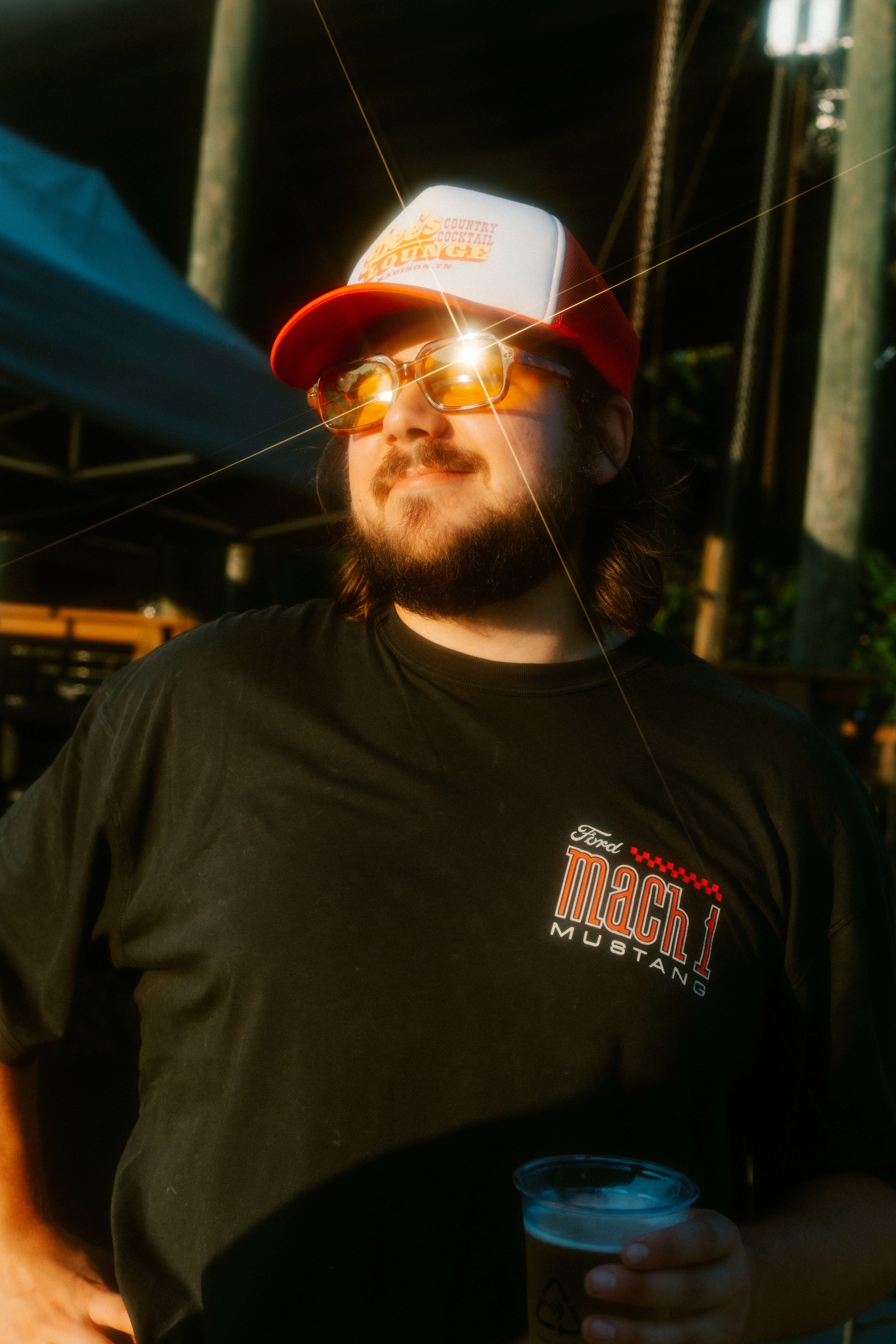 Man wearing sunglasses and a vintage hat stands confidently, holding a drink, as sunlight glimmers off his glasses. The background hints at a lively outdoor setting.