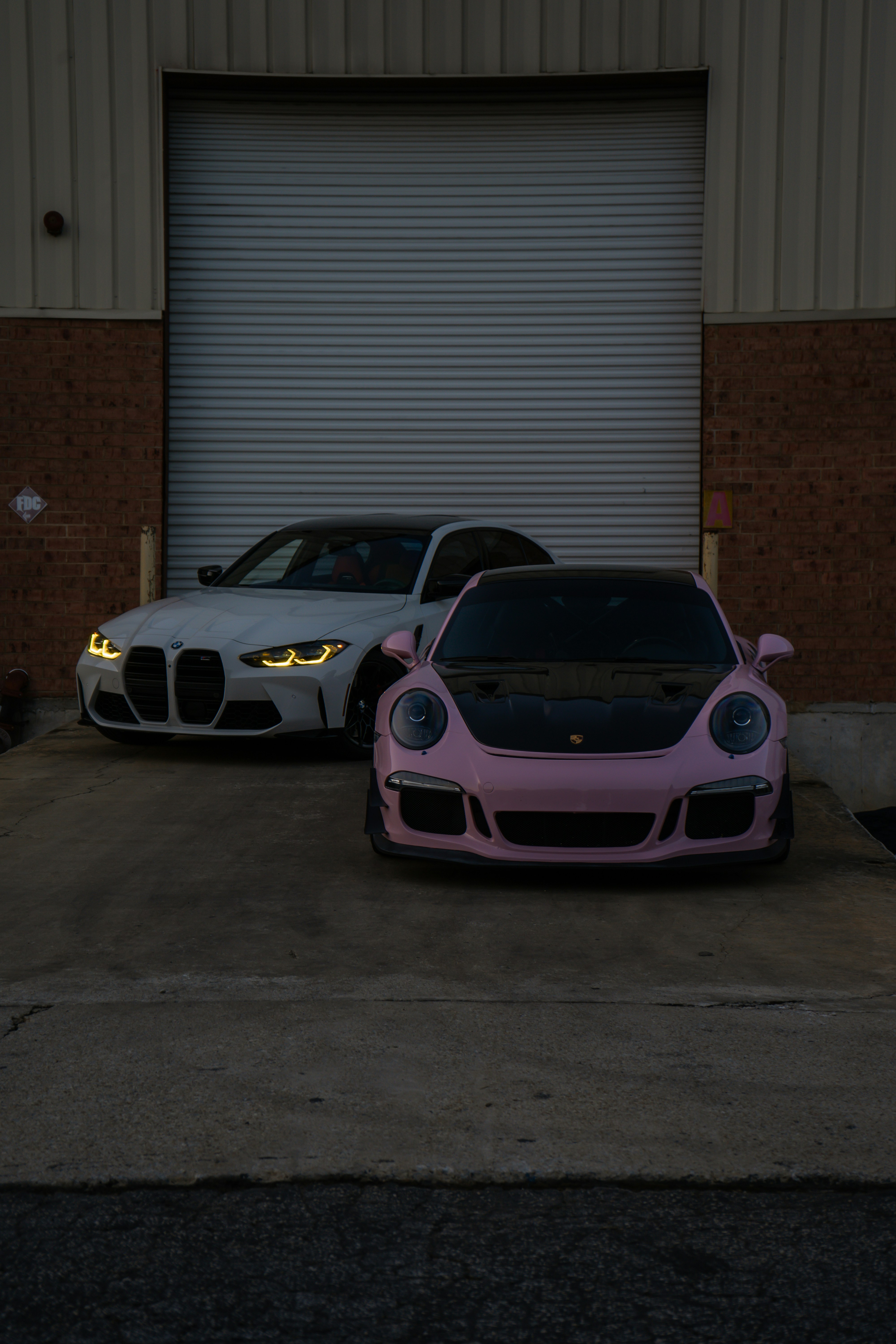 A sleek white BMW and a striking pink Porsche parked beside a closed garage door, showcasing their unique designs and colors in a dimly lit environment.