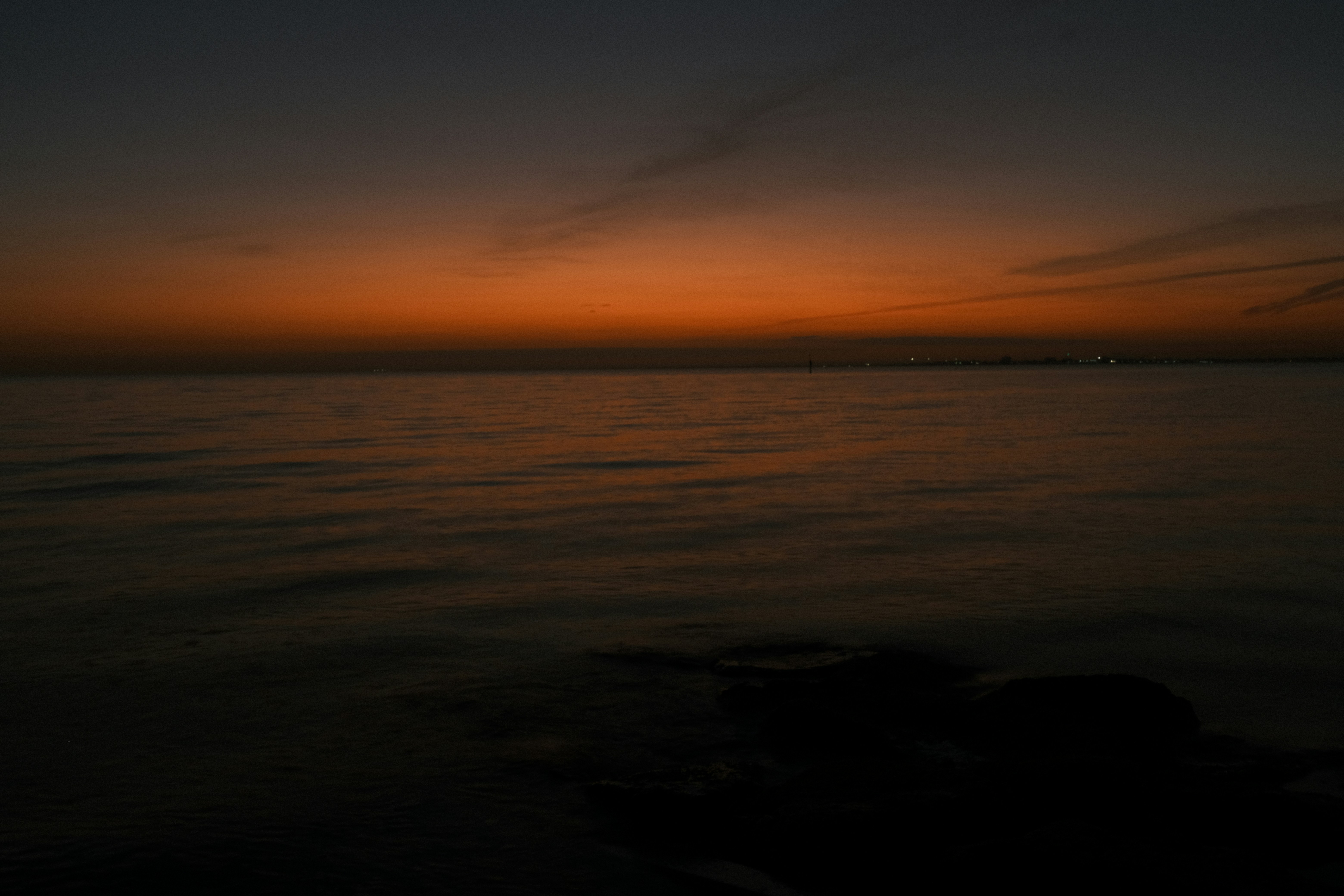 Serene twilight sky reflecting on calm ocean waters, with a faint outline of distant city lights on the horizon.