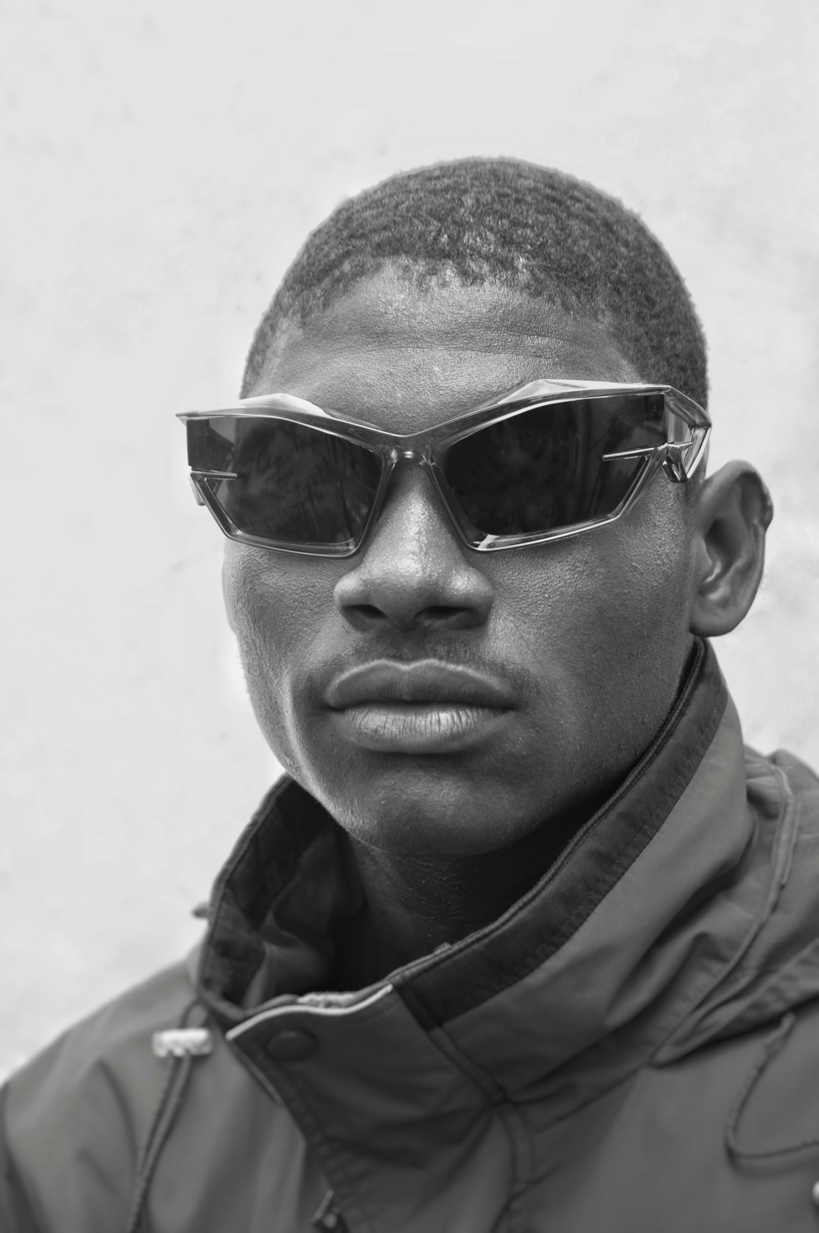 A young man exudes confidence in a stylish outfit, featuring striking sunglasses against a minimalist backdrop.