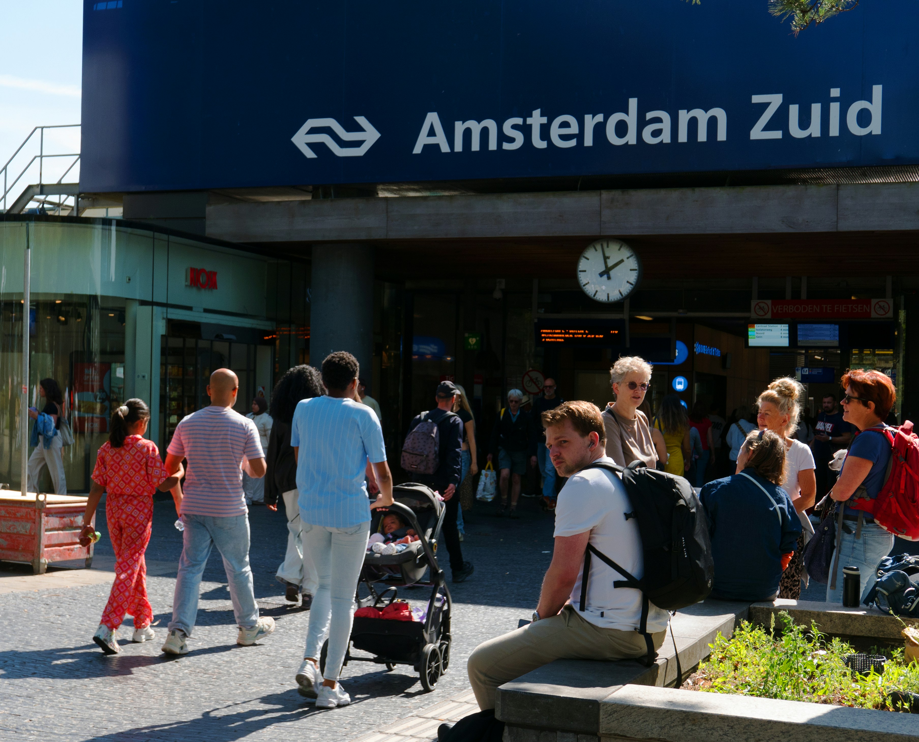 People are walking near the entrance of station Zuid in Amsterdam city on a sunny day with a lot of contrast. Free image. | People outside amsterdam zuid train station entrance