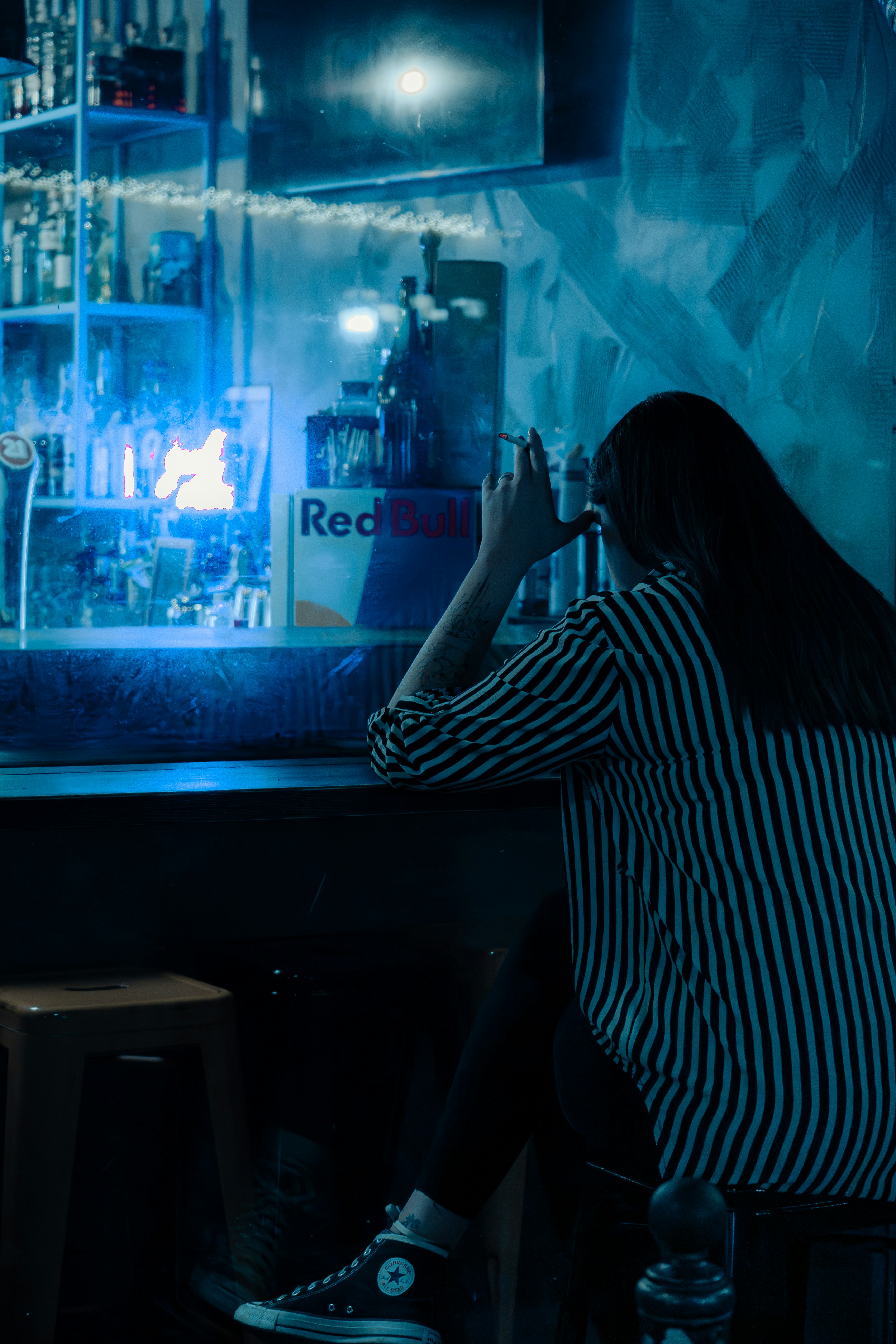 A solitary figure sits at a bar, framed by glowing neon lights and a backdrop of bottles, lost in thought. The atmosphere is intimate and moody.