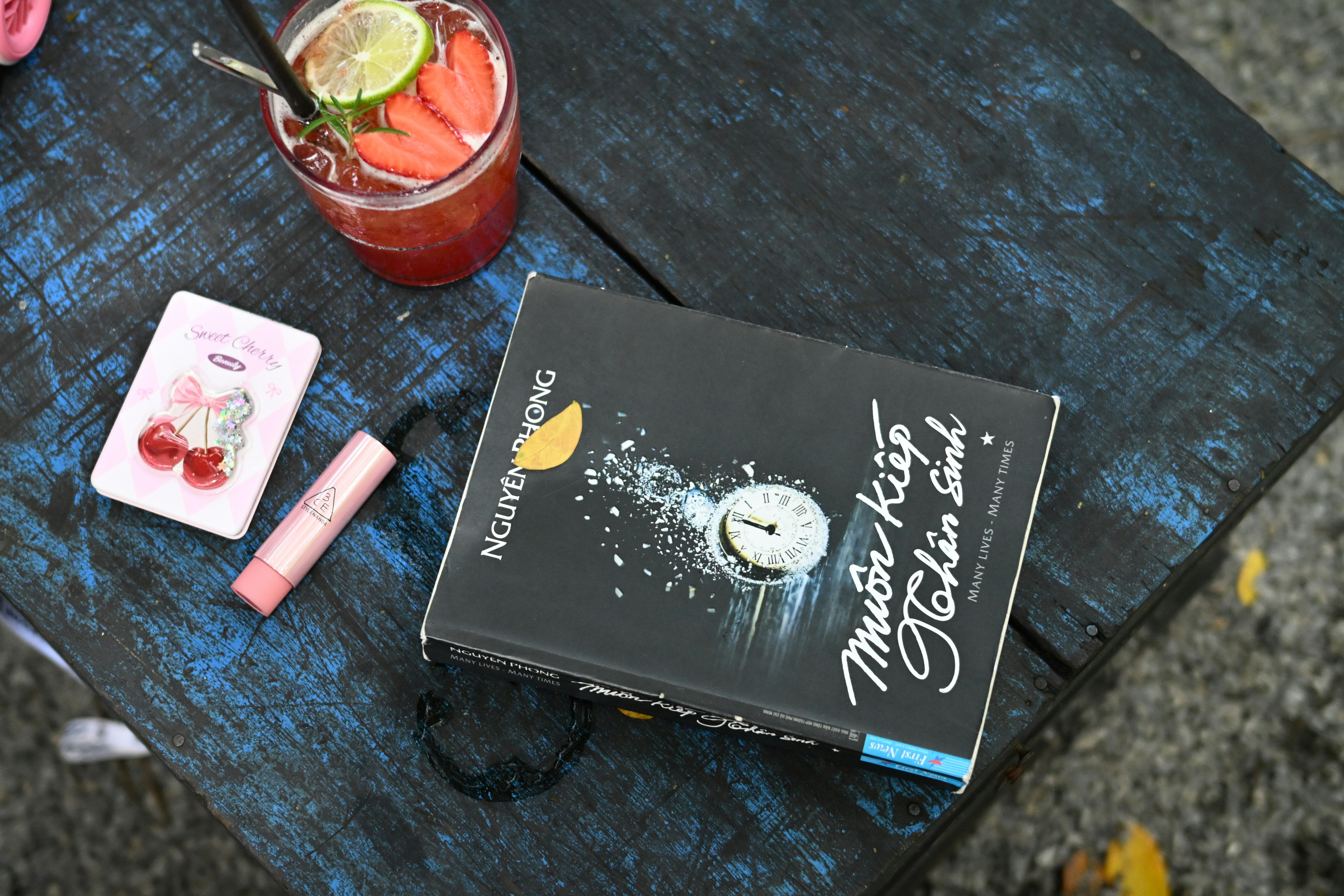 Open book titled 'Mưa Ký Tỏa Sáng' resting on a rustic blue table alongside a colorful drink with fruit garnishes and beauty items.