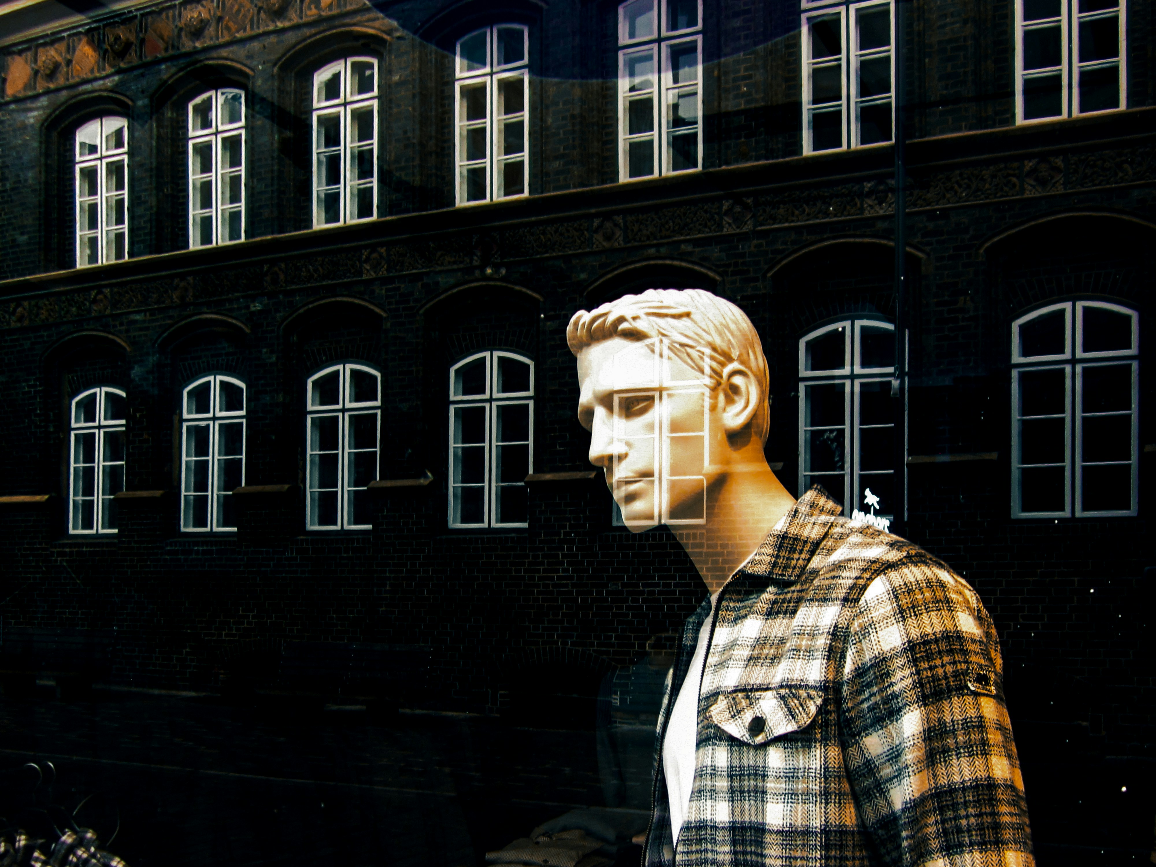 Urban reflection of a mannequin in Lübeck – street photography with a surreal twist. | Mannequin in plaid shirt against dark building.