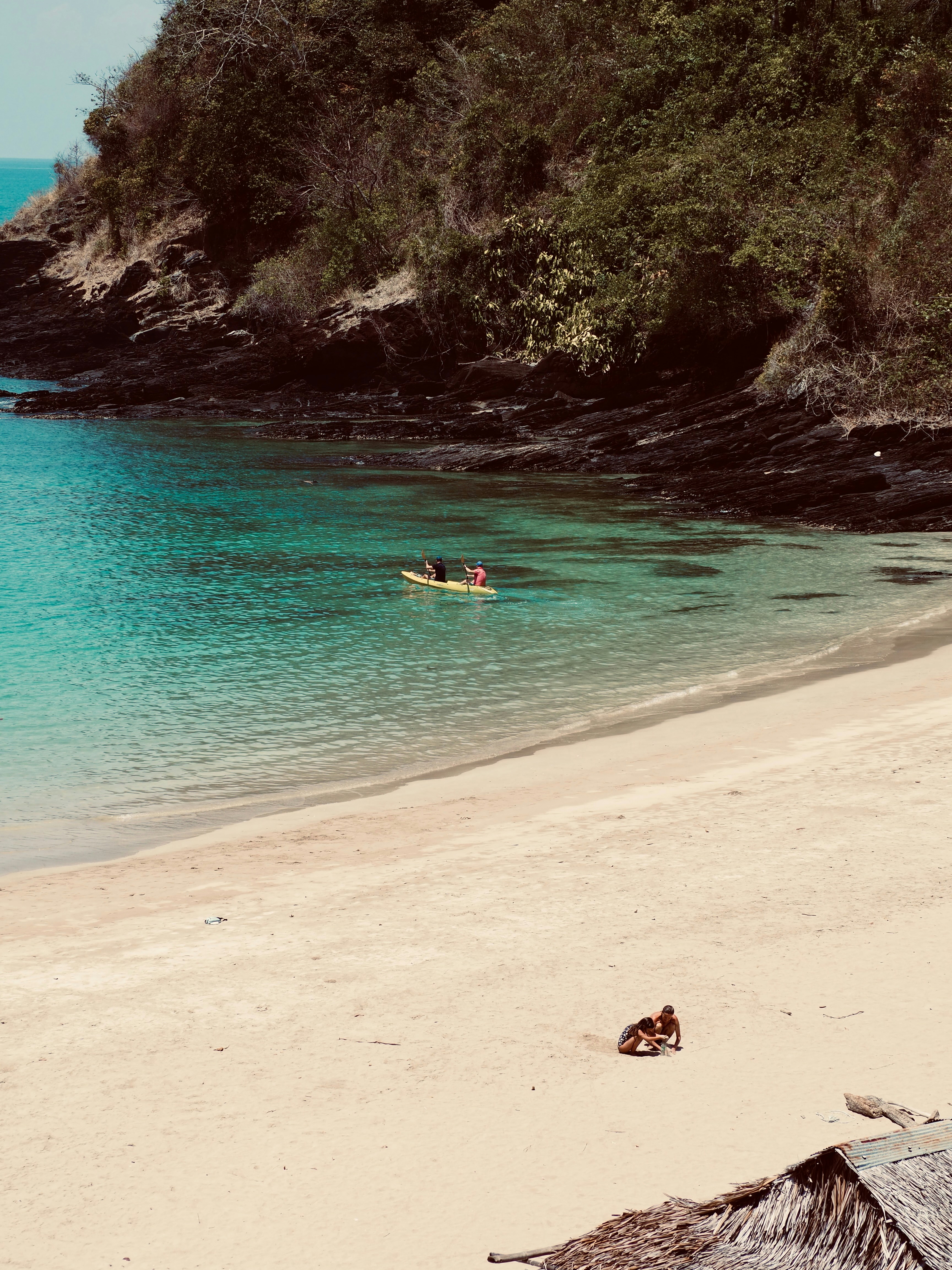 Two individuals kayaking in a serene cove, while another relaxes on the sandy beach under the warm sun.