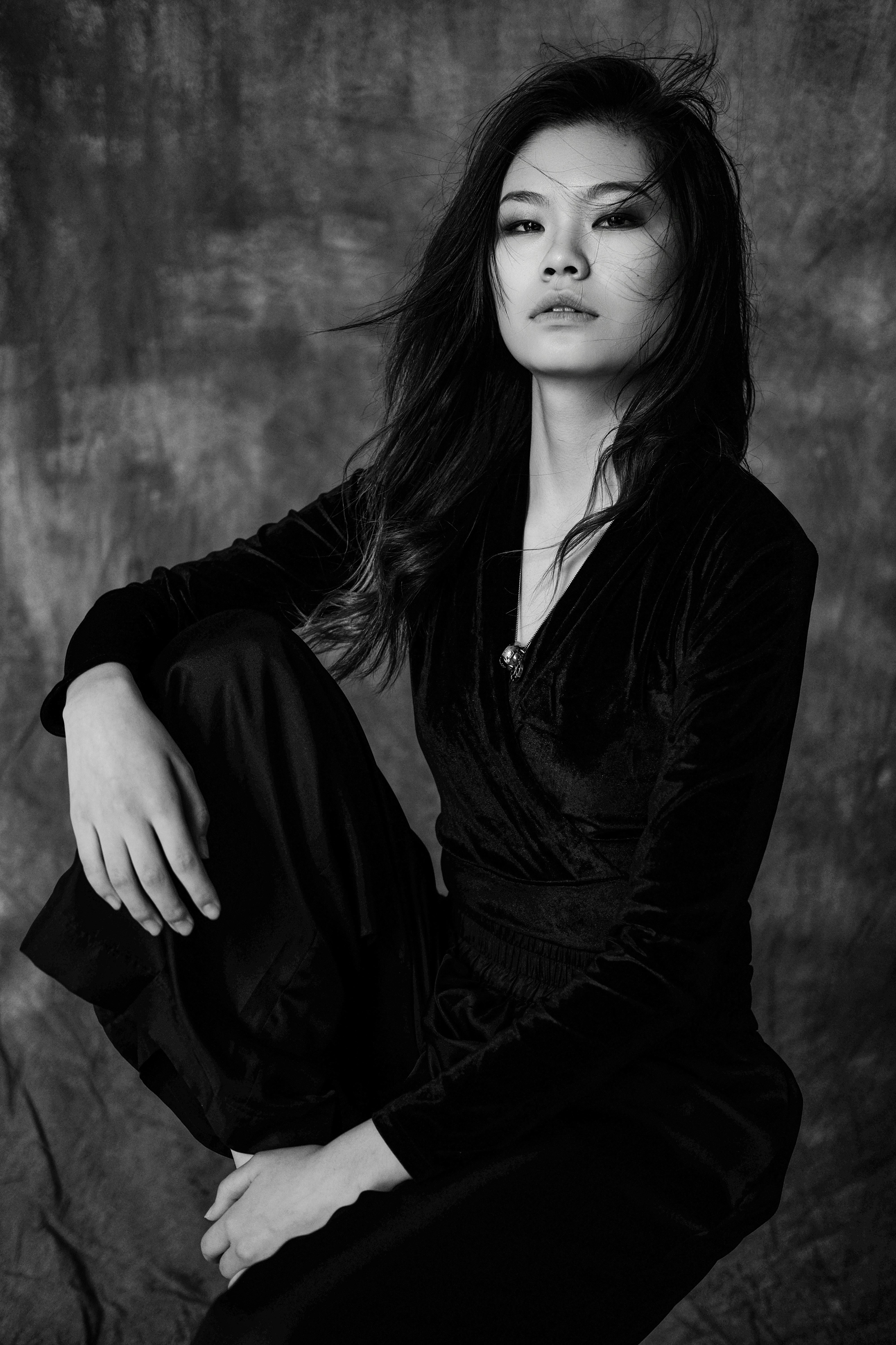 A poised young woman in a black velvet outfit, seated gracefully against a textured backdrop, exuding confidence and elegance.