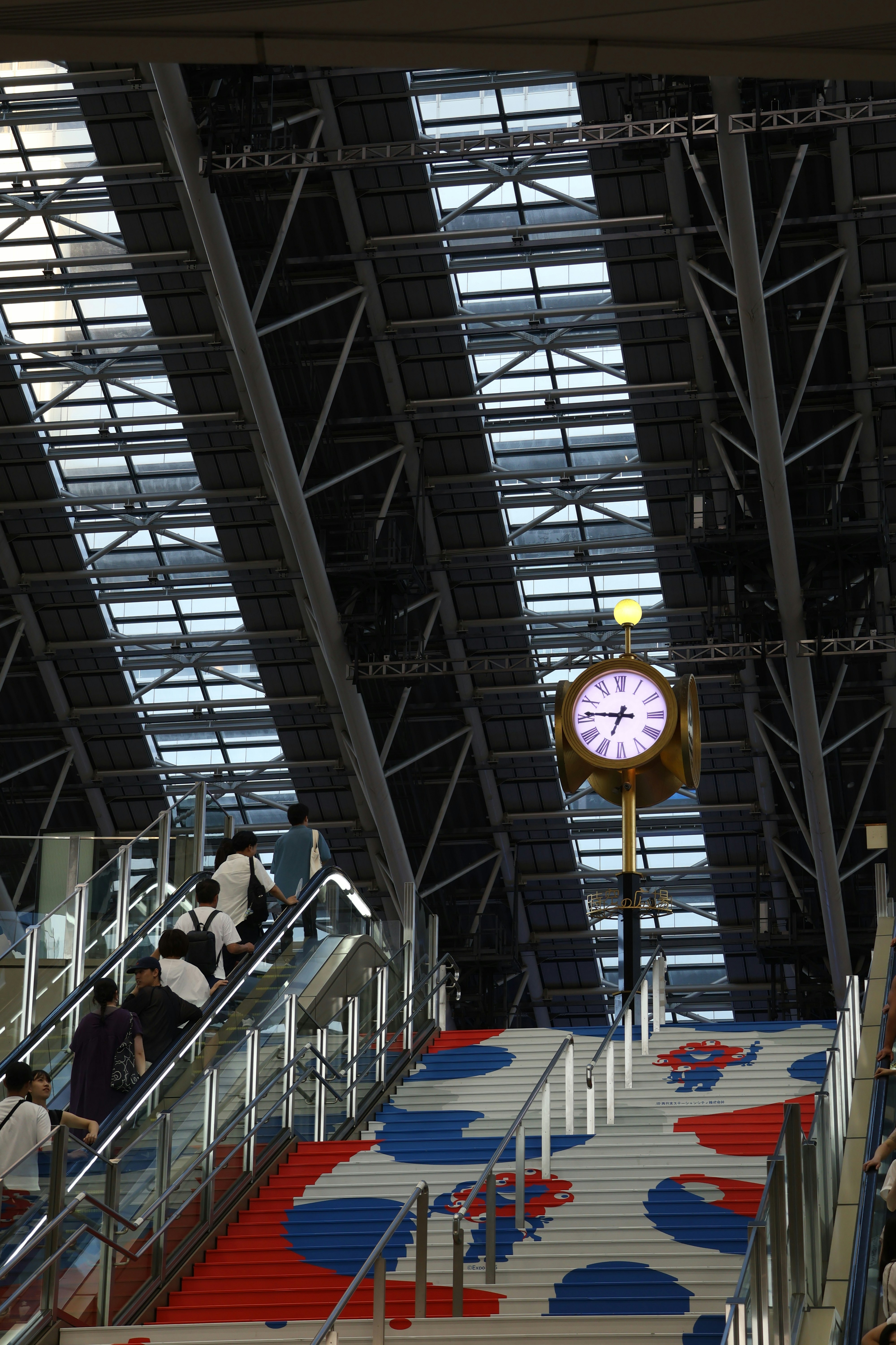 The Osaka expo is promoted all around the city, including the Osaka Station, a major transportation hub. | People ascend escalator beneath large clock