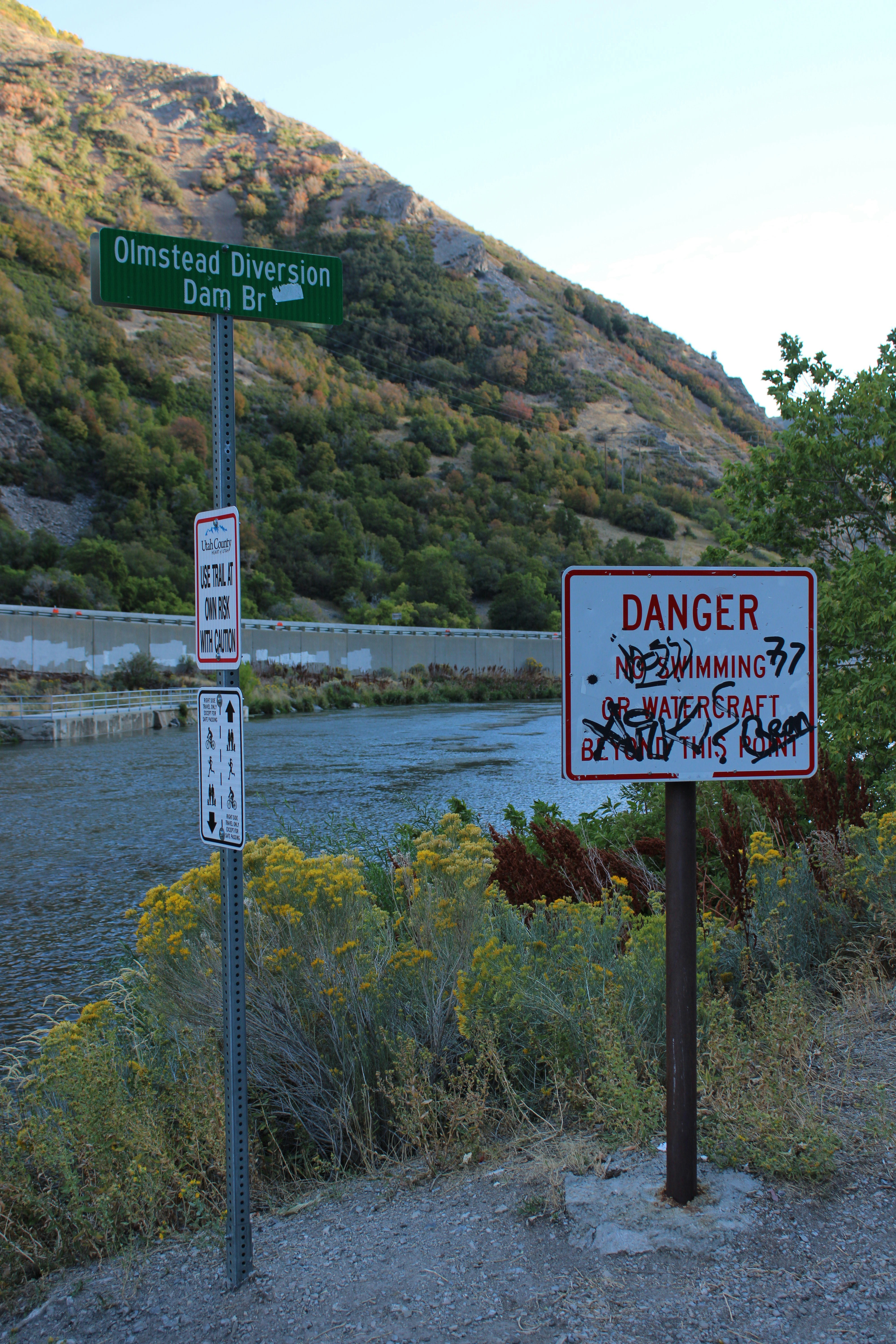 A warning sign near a river advises against swimming and watercraft, surrounded by lush vegetation and rocky terrain. The Olmstead Diversion Dam Bridge sign is visible in the background.