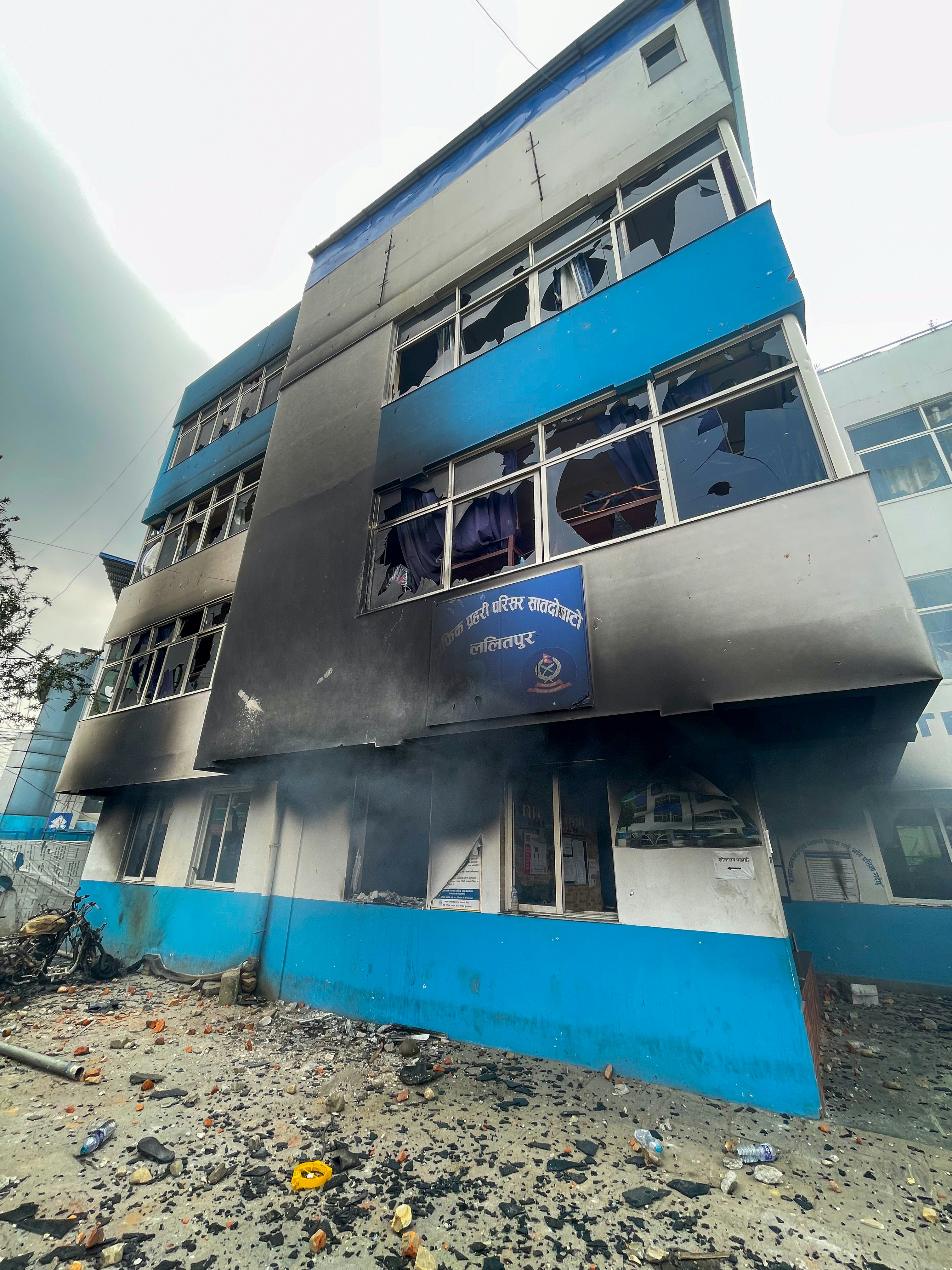 Wide shot of destroyed police office, broken windows, smoke Description: A wide-angle view of the Lalitpur police office shows shattered glass, burnt walls, and smoke rising after protesters attacked during the September 9 Nepal demonstration. The ground is littered with bricks and debris from the violent clashes. | Damaged building with smoke and debris after a fire