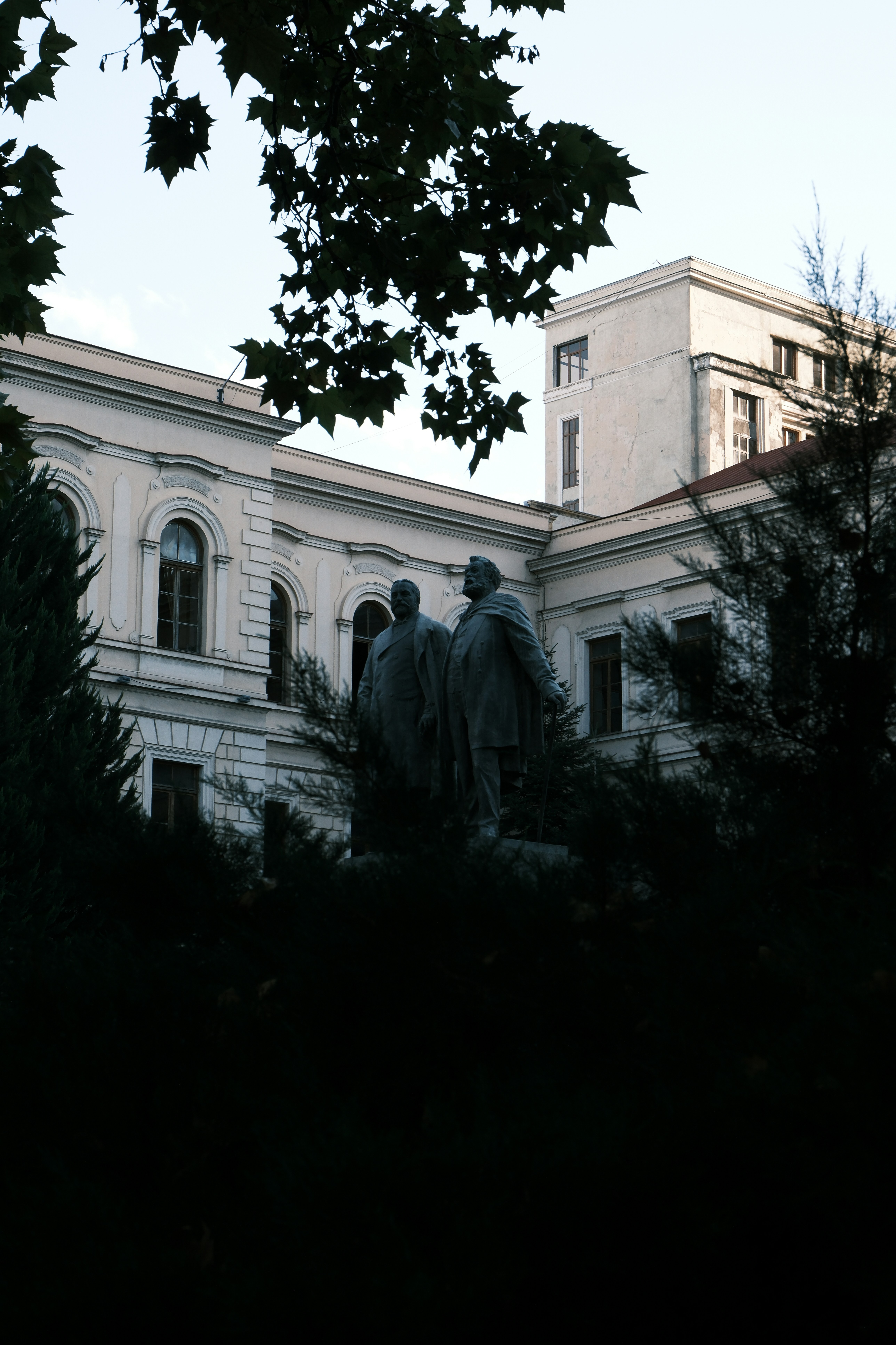 Two statues standing in a park, partially obscured by foliage, with a historic building in the background. The scene evokes a sense of reflection and nostalgia.