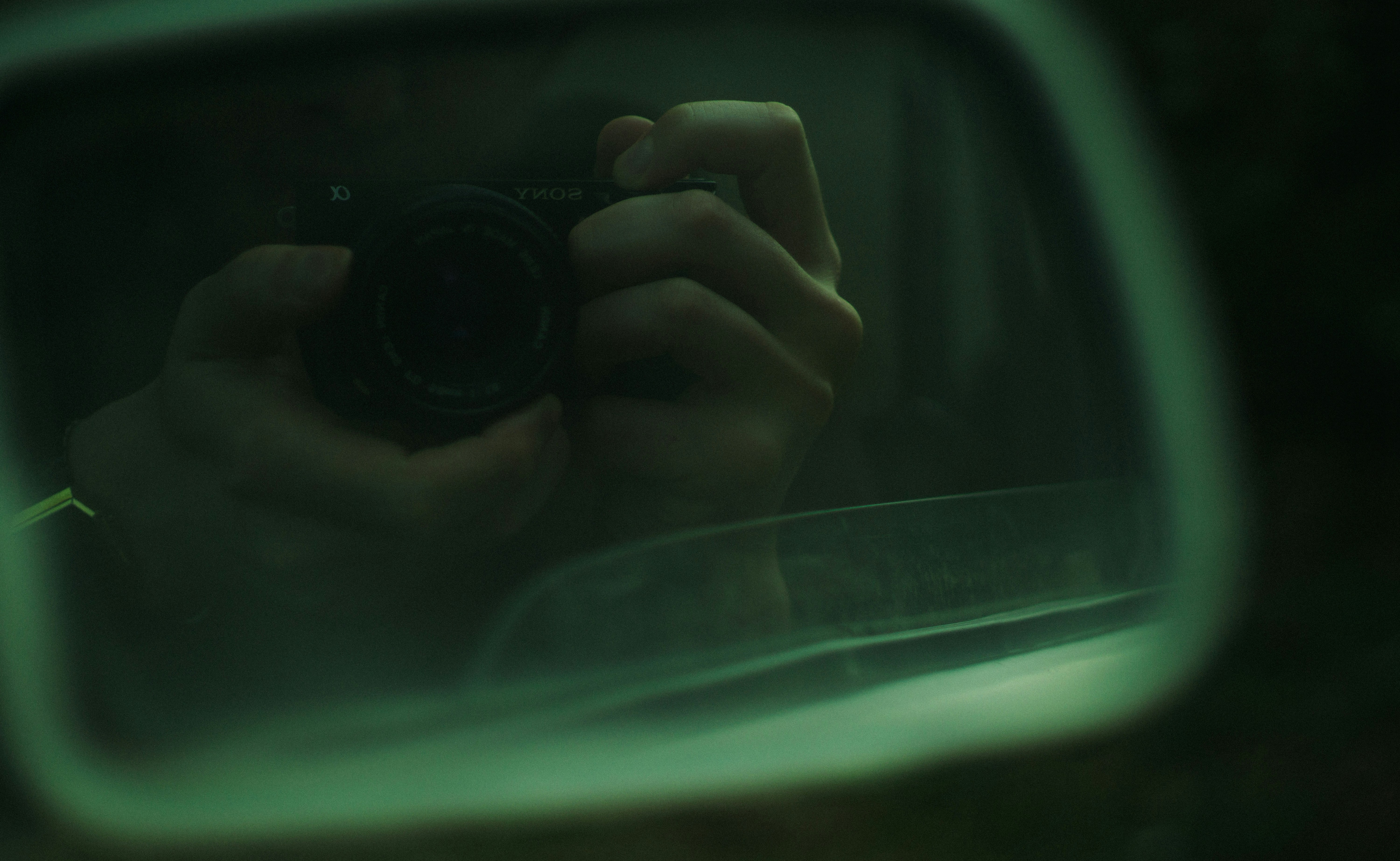 Reflection of a photographer holding a camera in a car side mirror, surrounded by a blurred natural backdrop.