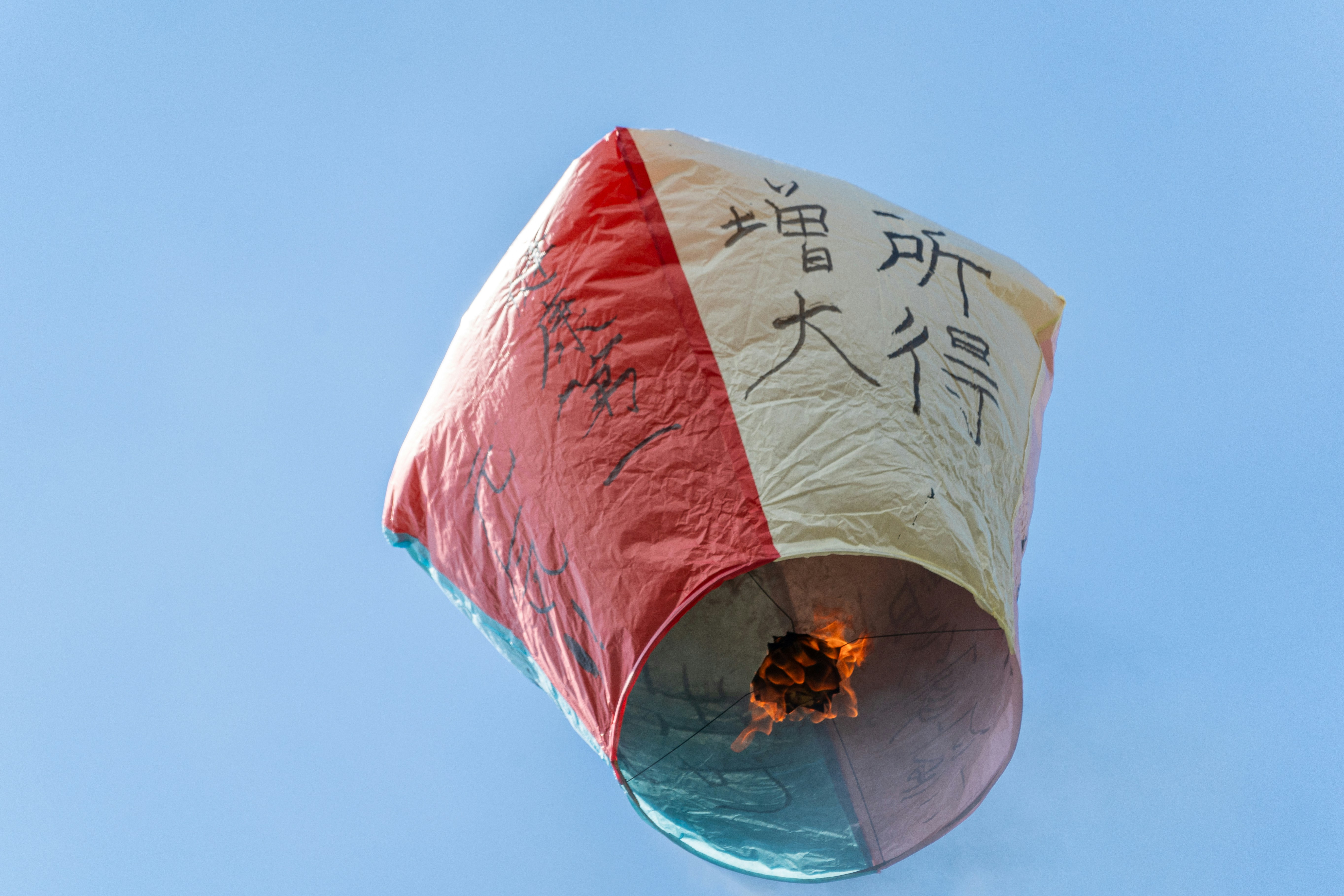 Colorful sky lantern drifting against a clear blue sky, featuring intricate designs and calligraphy. The lantern's flame illuminates its interior as it rises.