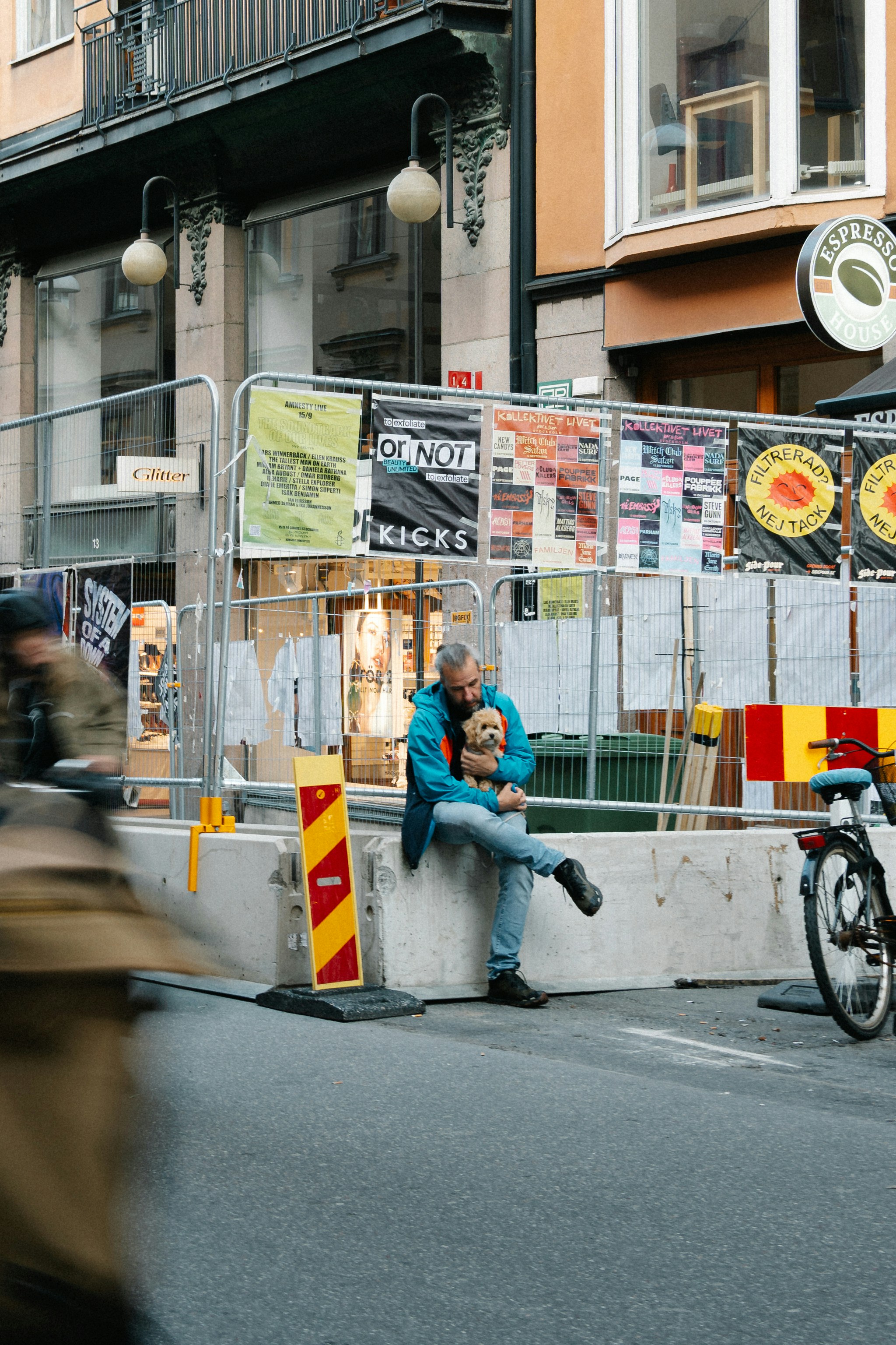 A man sits on a construction barrier, cradling a small dog, surrounded by busy street activity and colorful posters. The scene captures a moment of calm in a bustling urban environment.