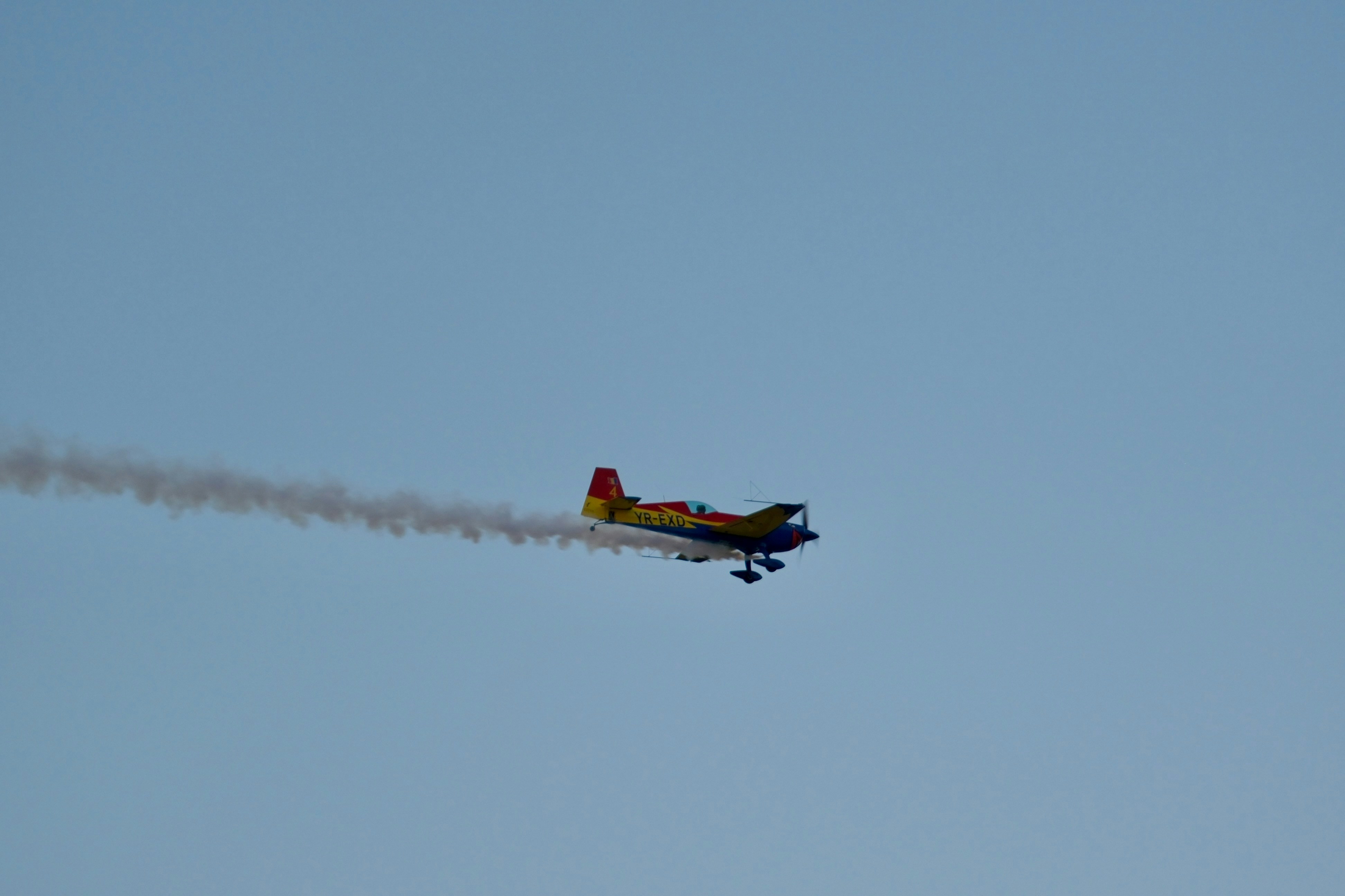Colorful stunt plane performing aerial maneuvers against a clear blue sky, leaving a trail of smoke behind.