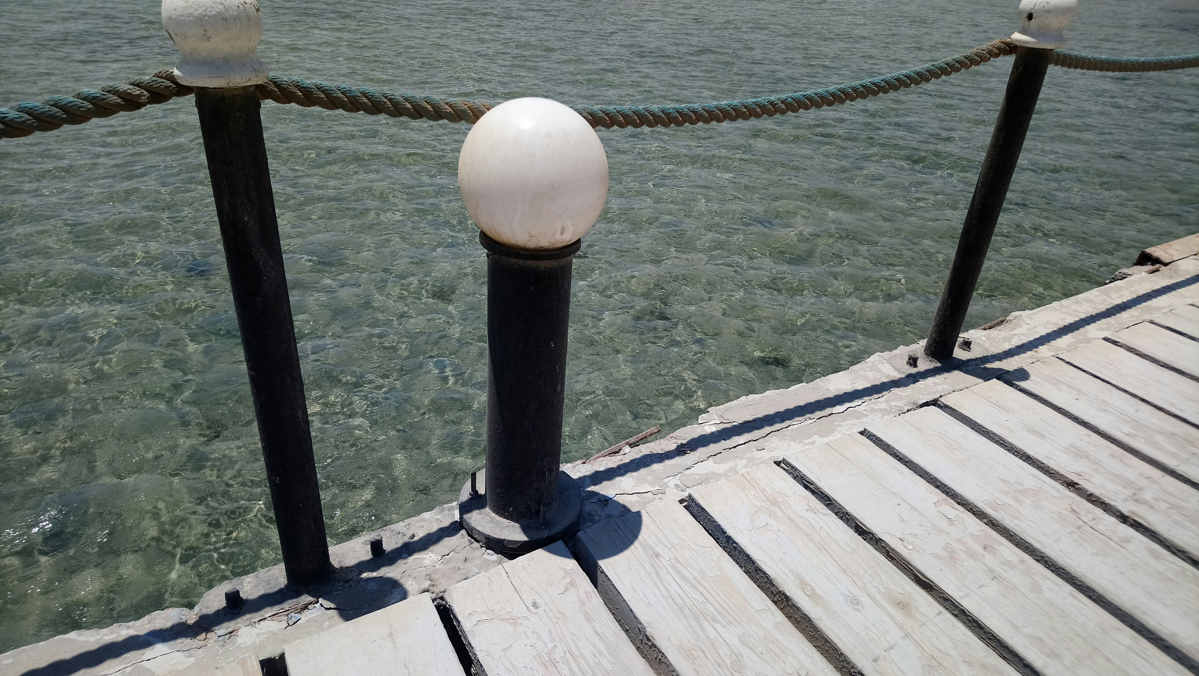 A long wooden pier that extends into the sea with posts on which lanterns are placed. | Wooden pier with rope railing and globe lights over water