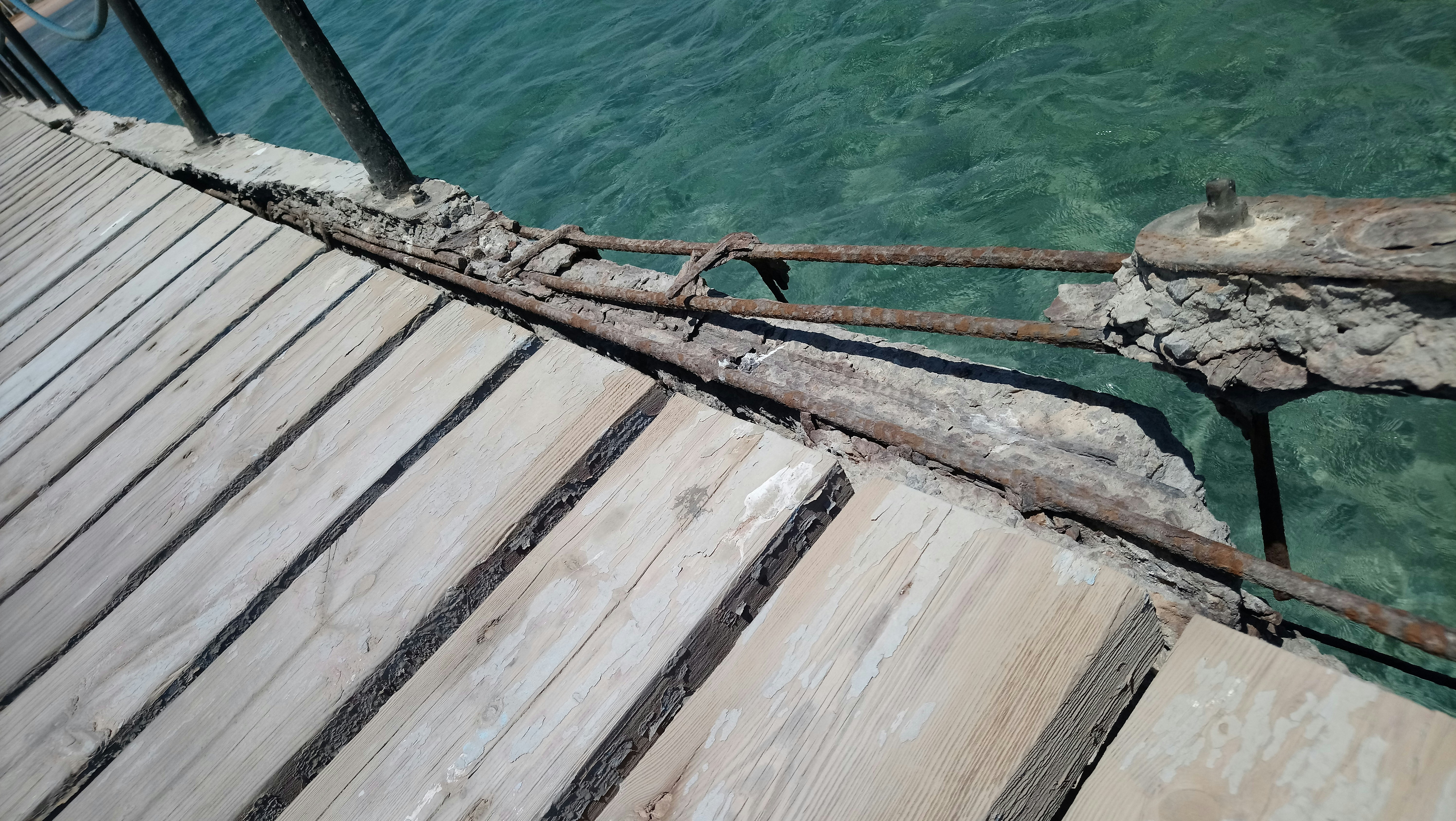 An old sea wooden pier. It is almost distroyed. | Wooden walkway over clear turquoise water with rusty railing.