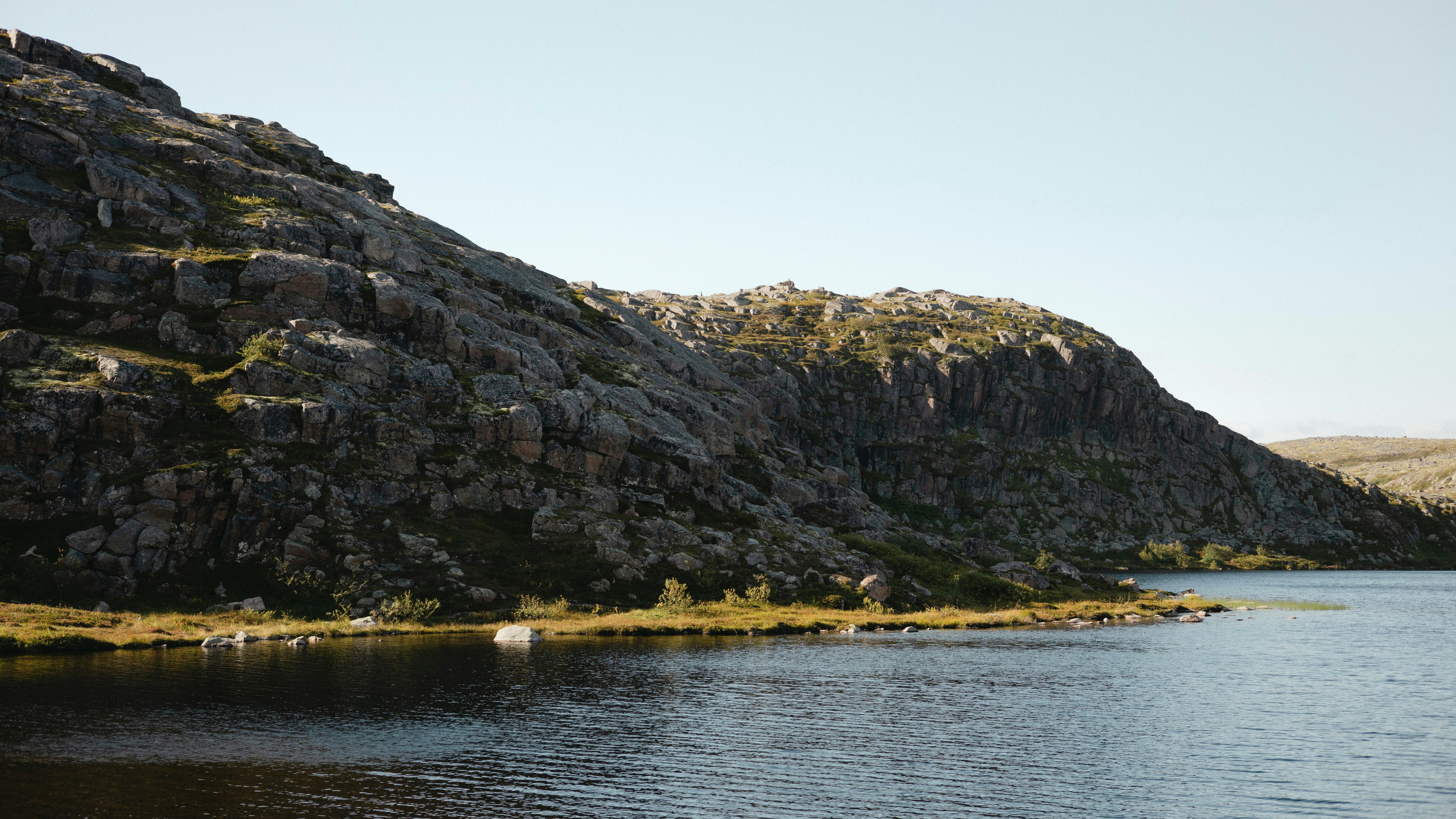 Lush, rugged hills reflect in a serene lake under a clear sky, showcasing the tranquil beauty of nature's formations.