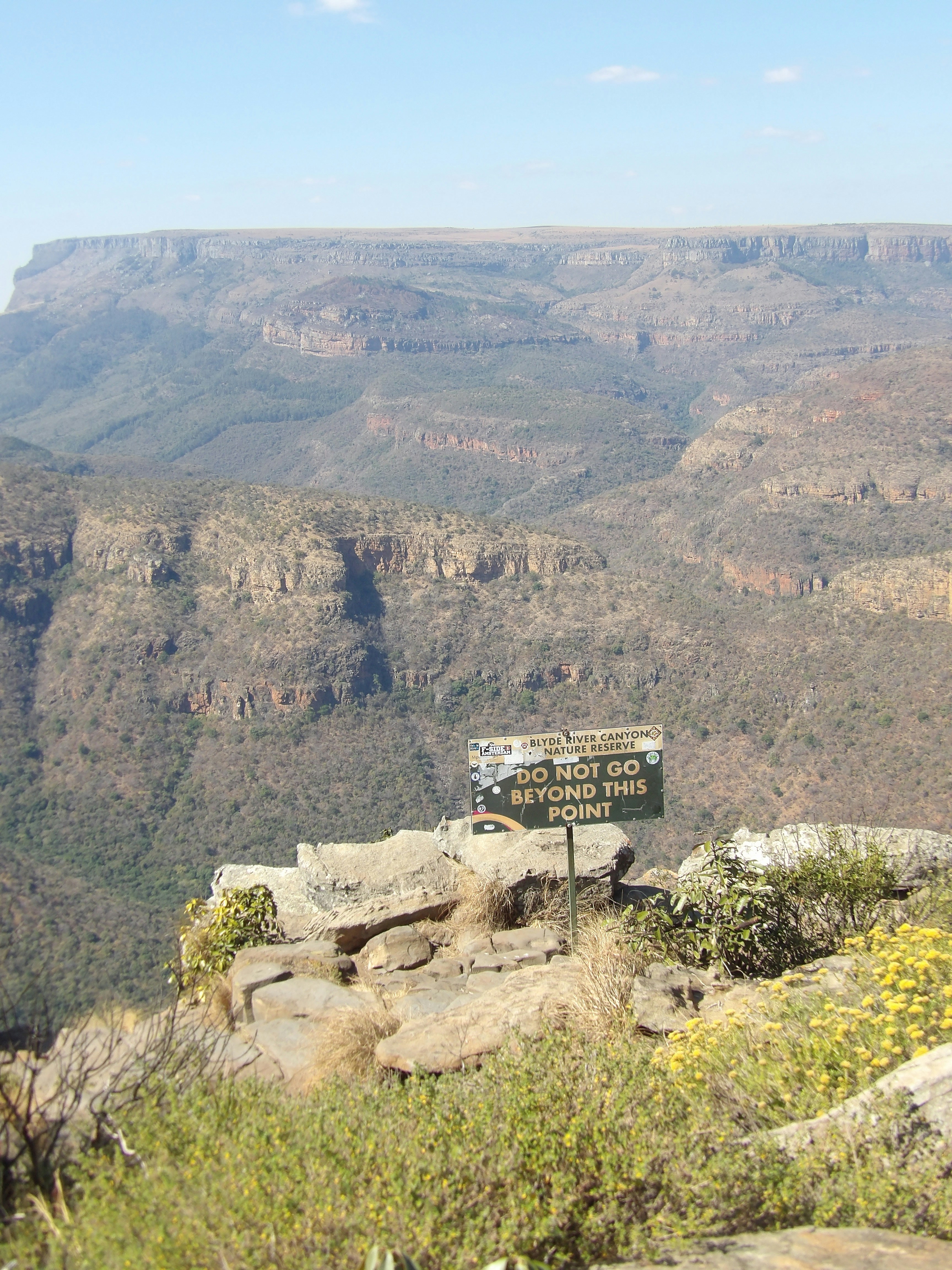 Warning sign at the edge of a canyon overlooking rugged terrain and distant mountains. The sign advises against proceeding beyond the marked point.