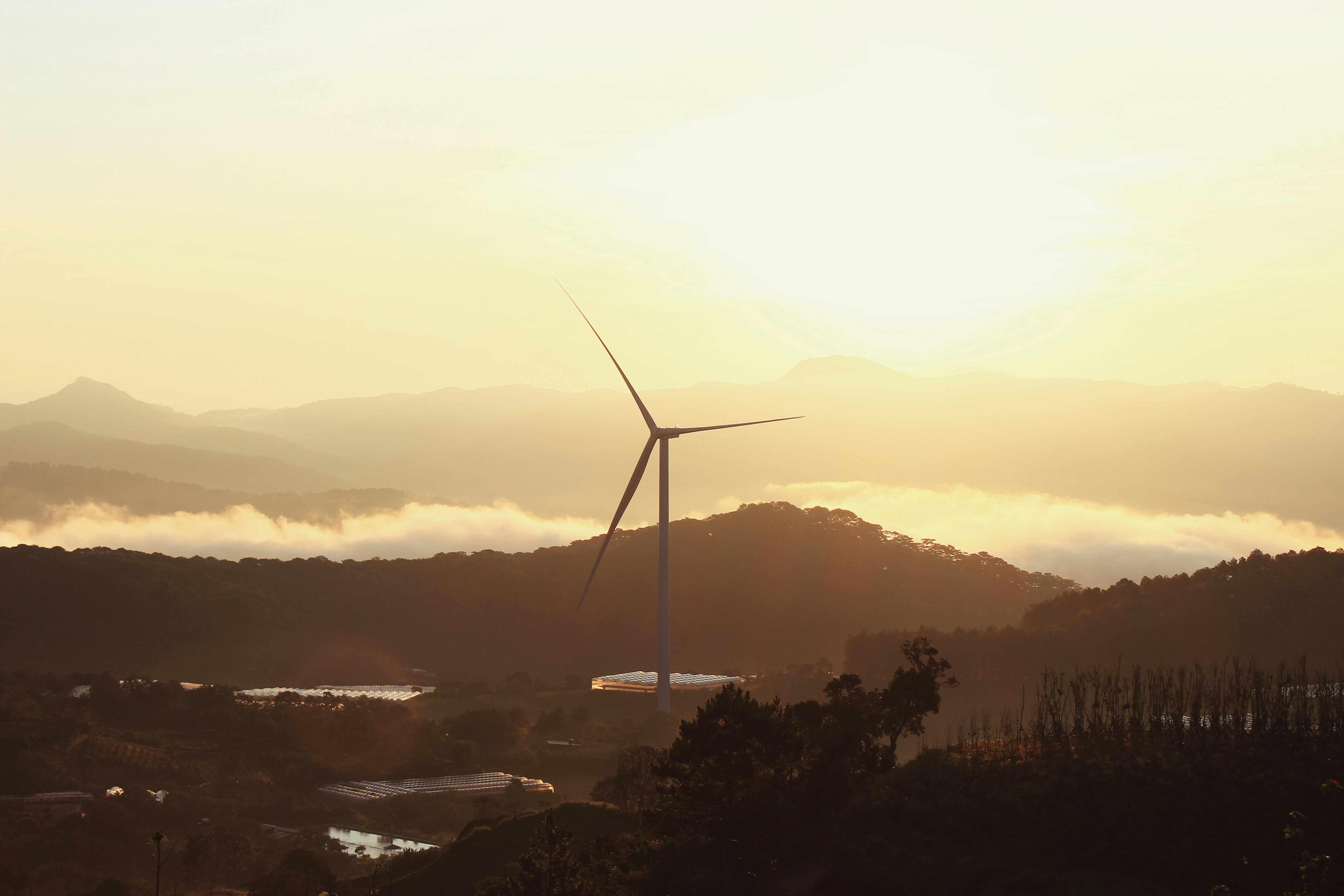 Wind turbine silhouetted against a hazy sunrise over rolling hills.