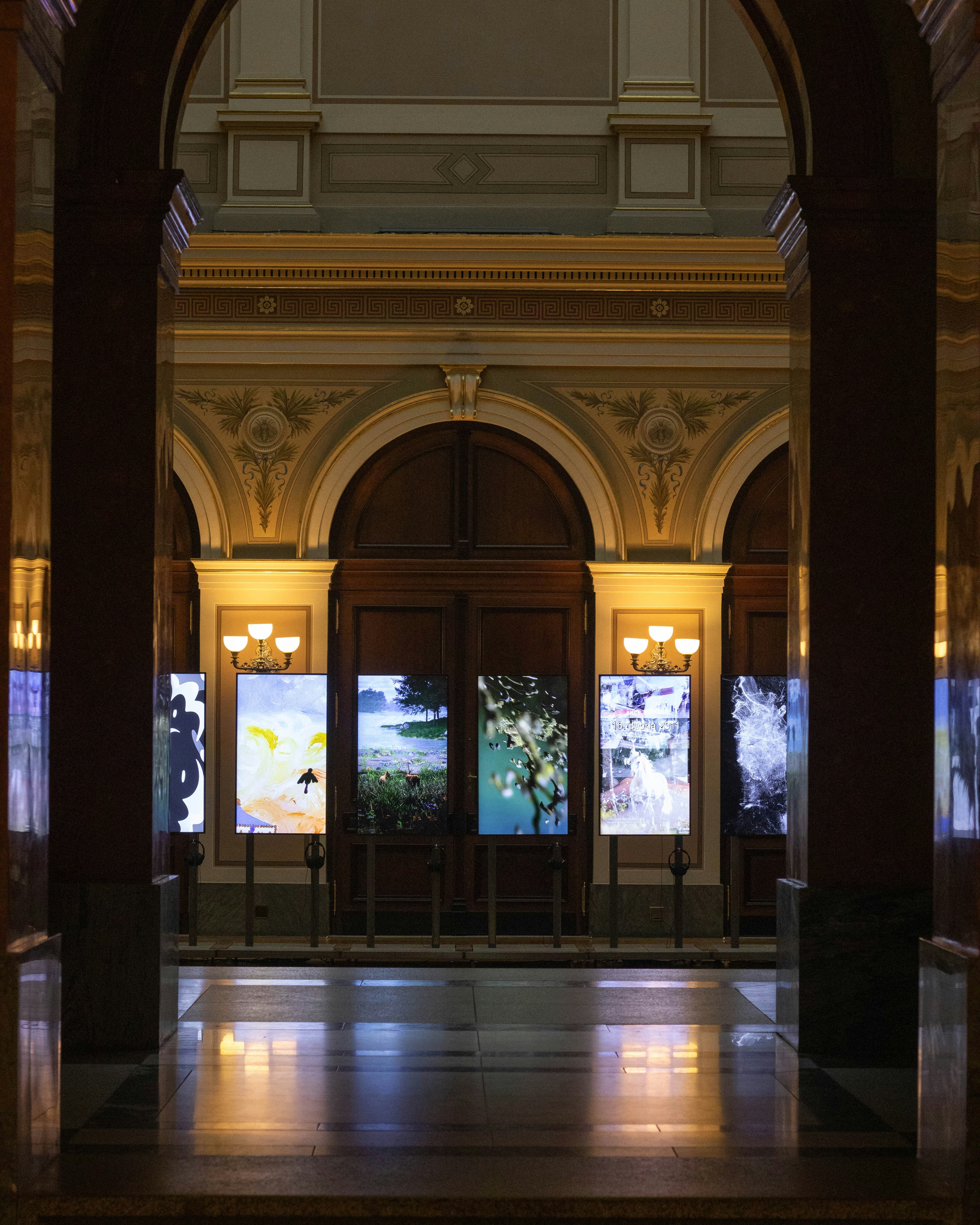 Art exhibition in a grand hall with illuminated paintings.