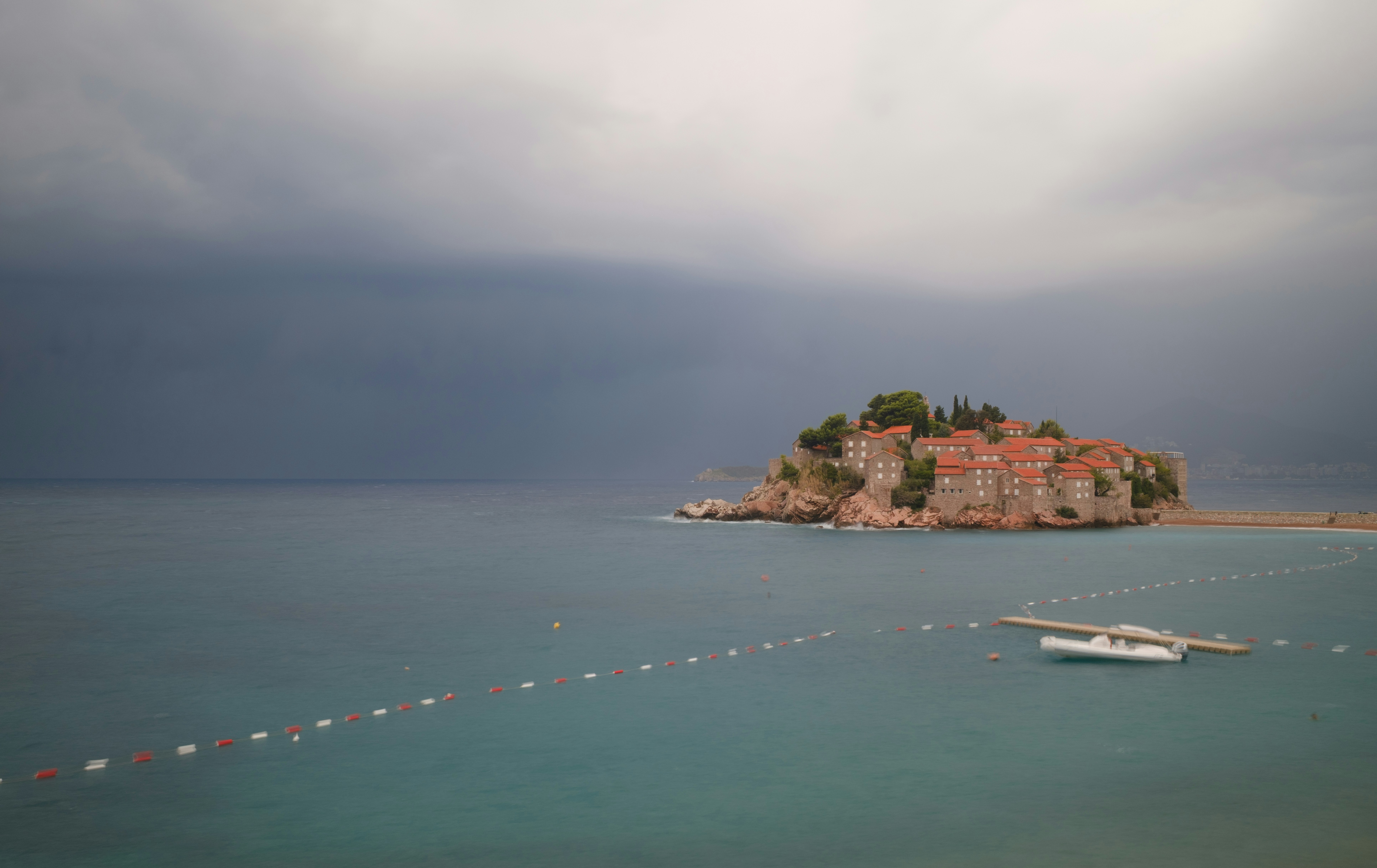 A picturesque island village with terracotta roofs sits against a backdrop of darkening skies and calm turquoise waters.