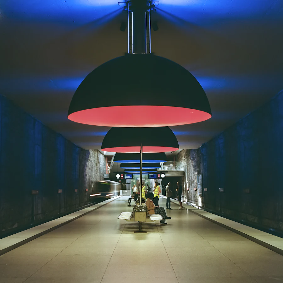 A long subway corridor lined with oversized black dome lamps glowing pink underneath, with a few people seated and standing near the center. Blue light washes the ceiling and walls toward the far end of the platform.