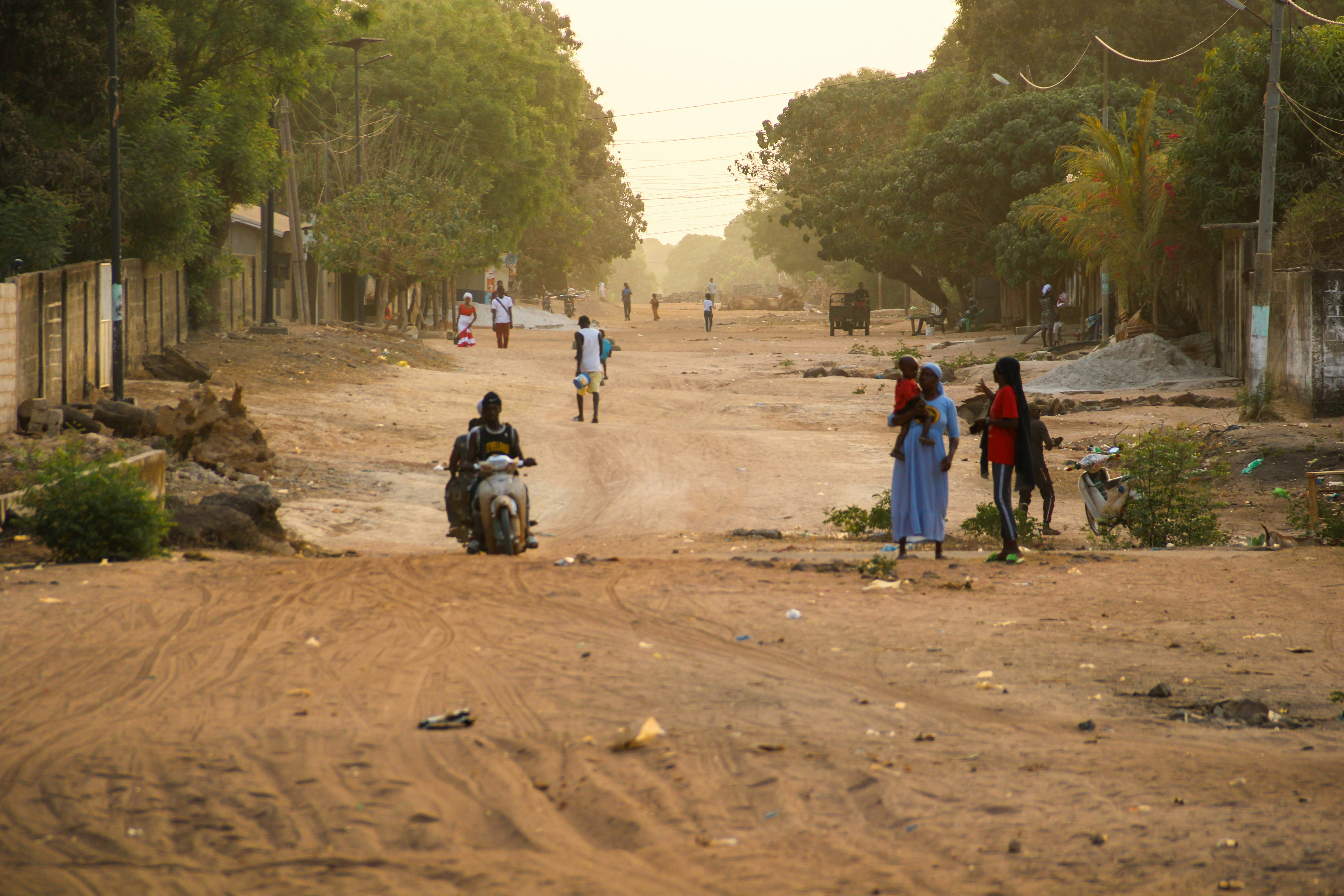 Morning traffic in Bignona, Senegal. | People walking on a dusty road in a village.