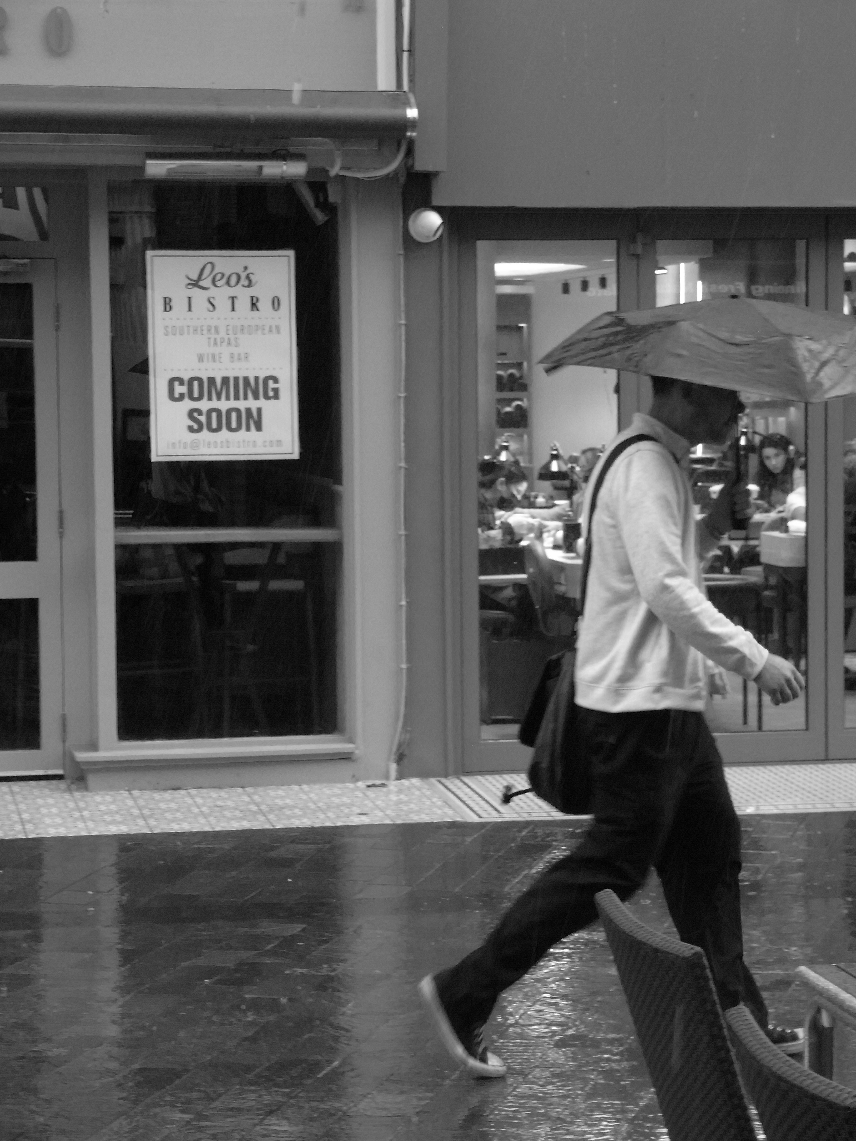 A person walks with an umbrella past a bistro under rainy skies, showcasing the hustle of urban life. The scene is captured in black and white, emphasizing the contrast and mood.