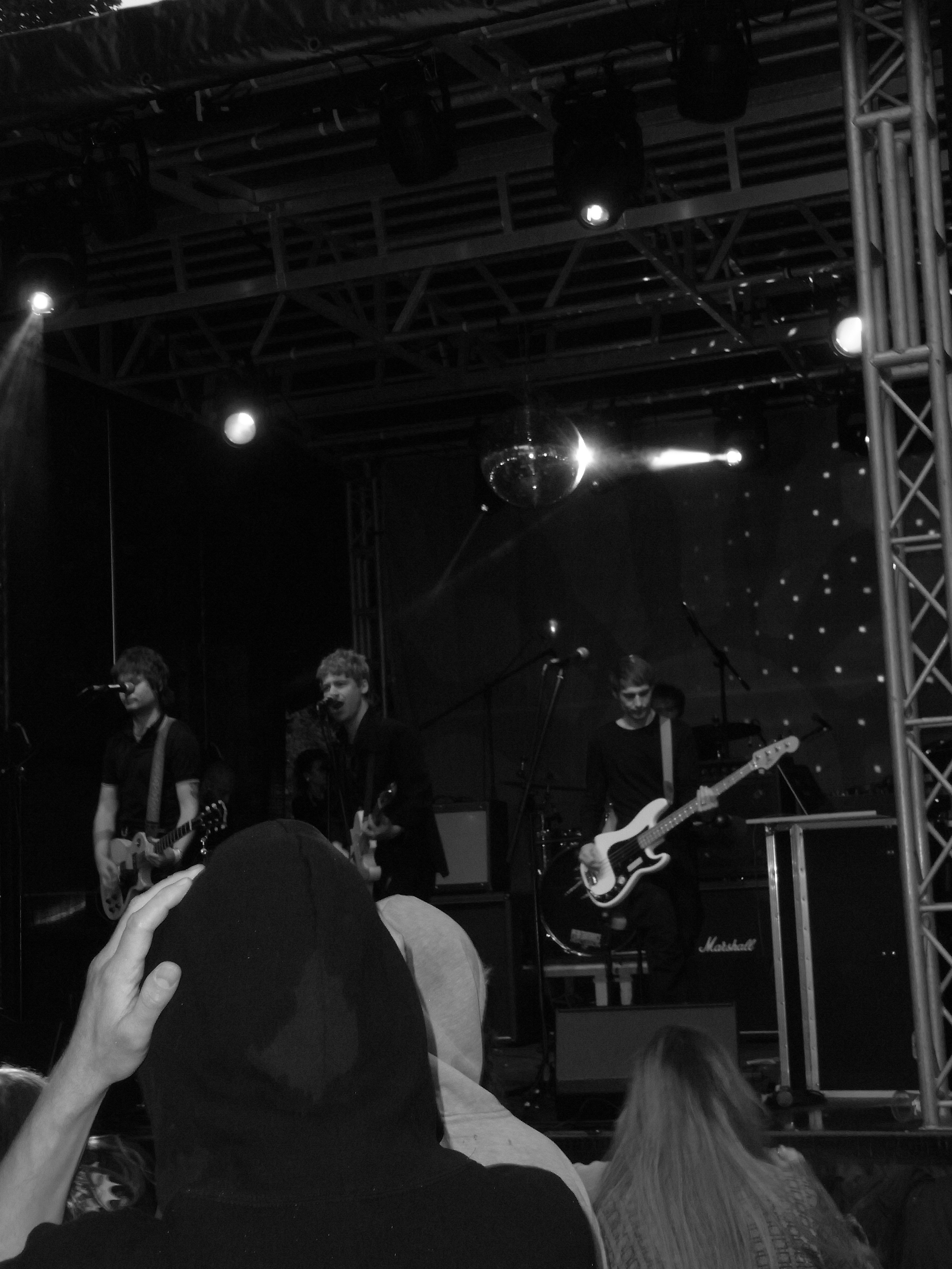 Three musicians performing on stage under dramatic lighting, with a captivated audience in the foreground. The scene captures the energy of a live concert.