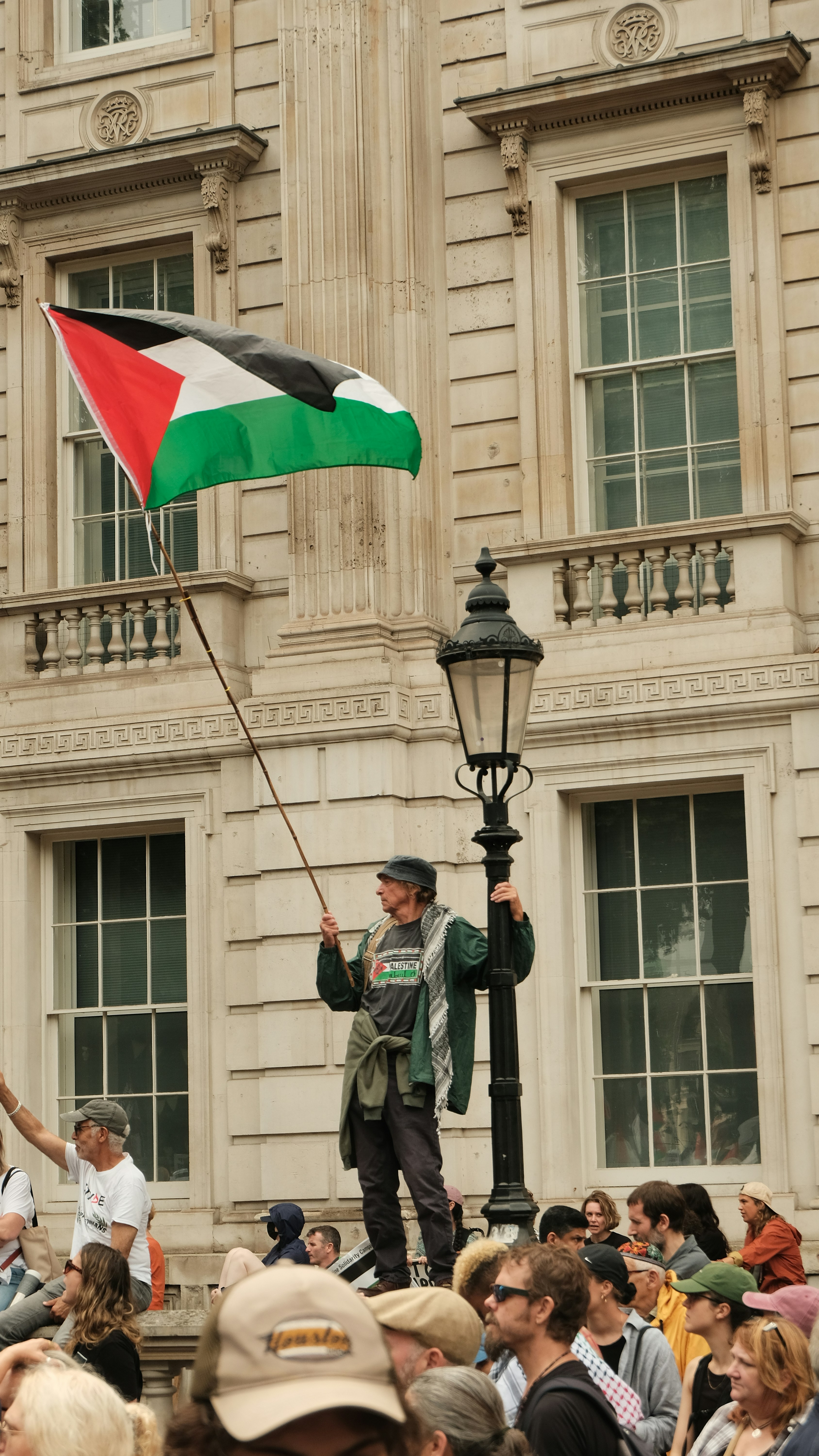 Activist holding a Palestinian flag amidst a crowd during a demonstration in front of a historic building.